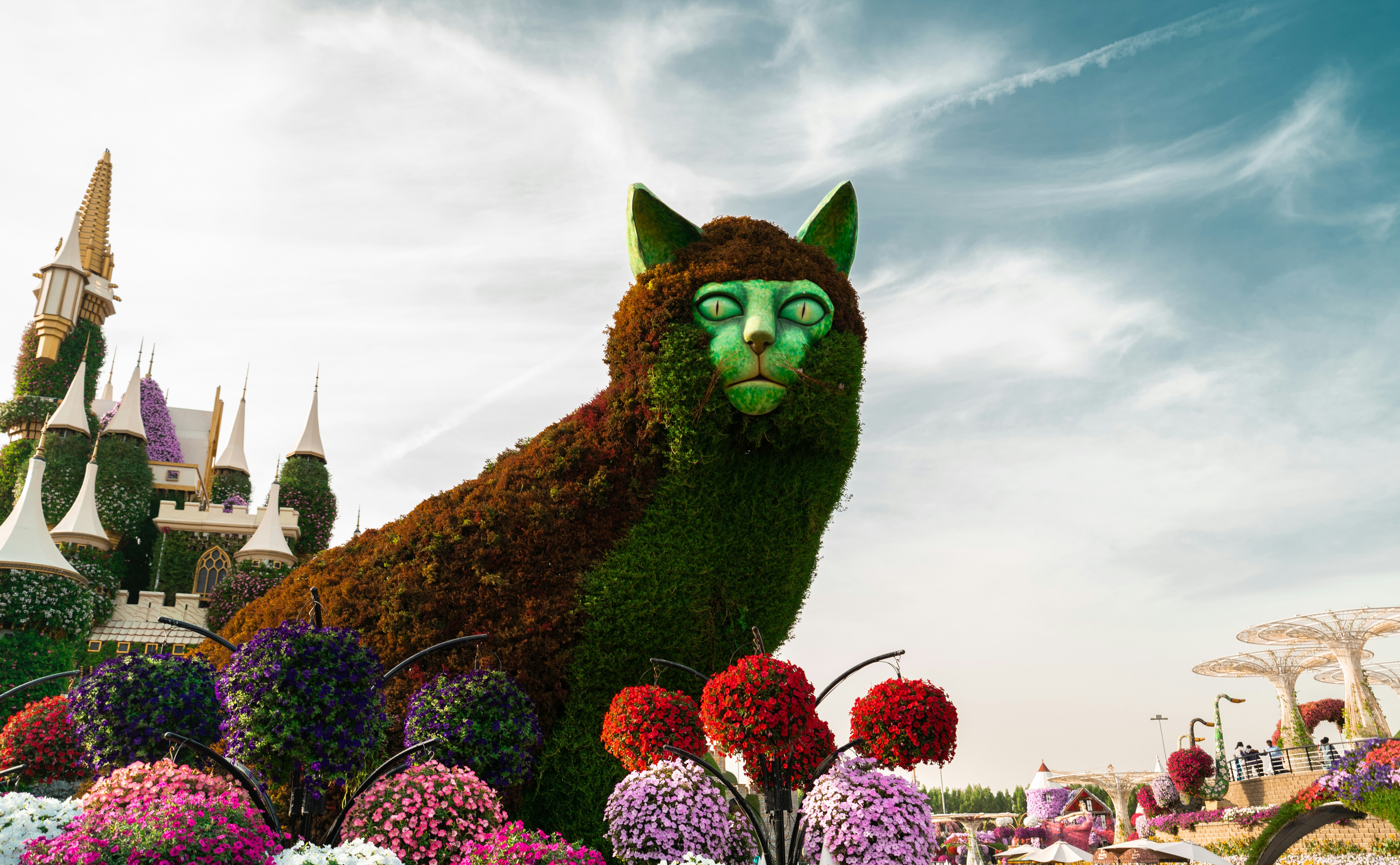 A colossal topiary cat with a vibrant green hue stands majestically among colorful flower arrangements, set against a whimsical castle backdrop.