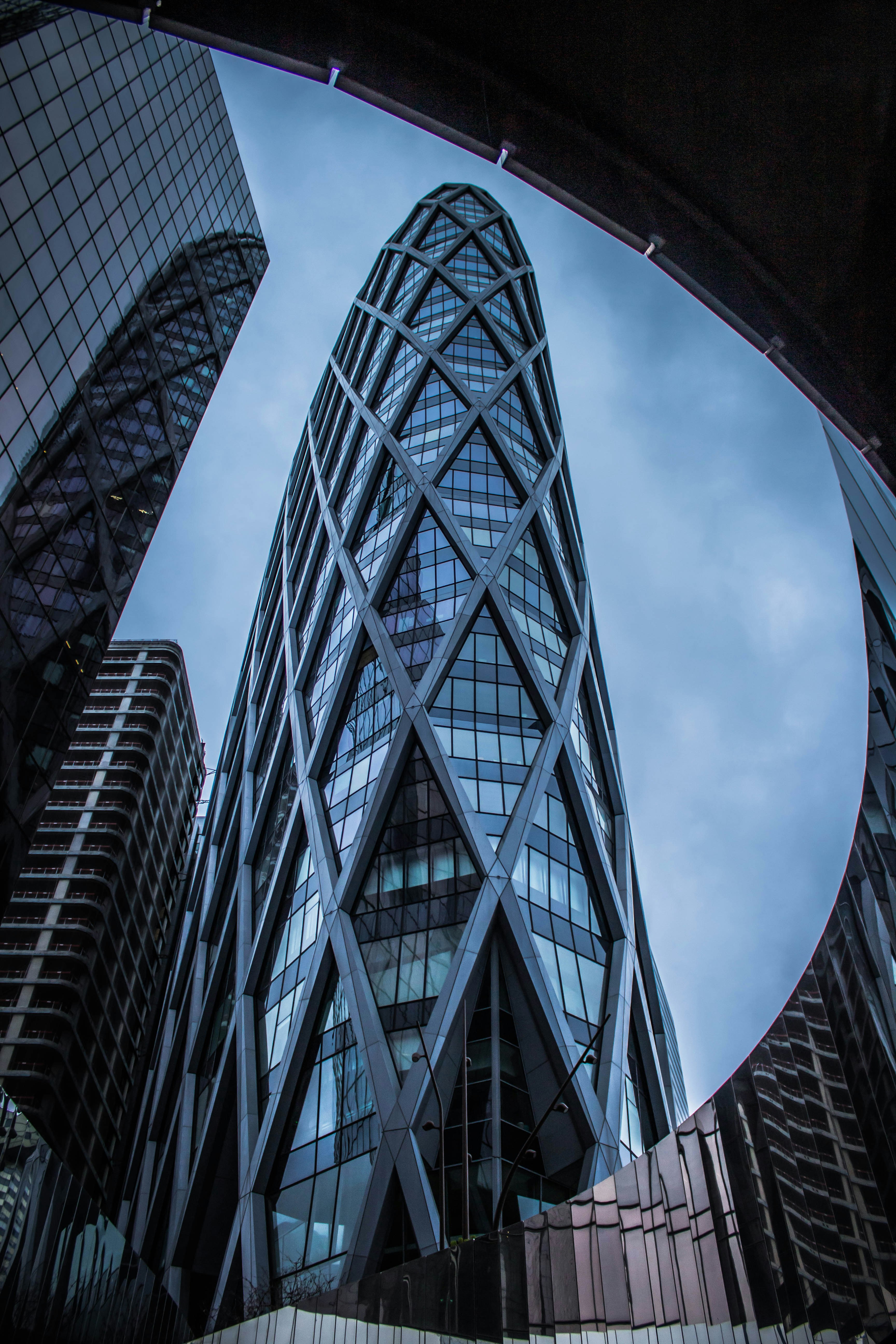 A modern skyscraper with a distinctive diamond-patterned facade reaches towards a cloudy sky, framed by surrounding buildings. The architectural design emphasizes height and symmetry.