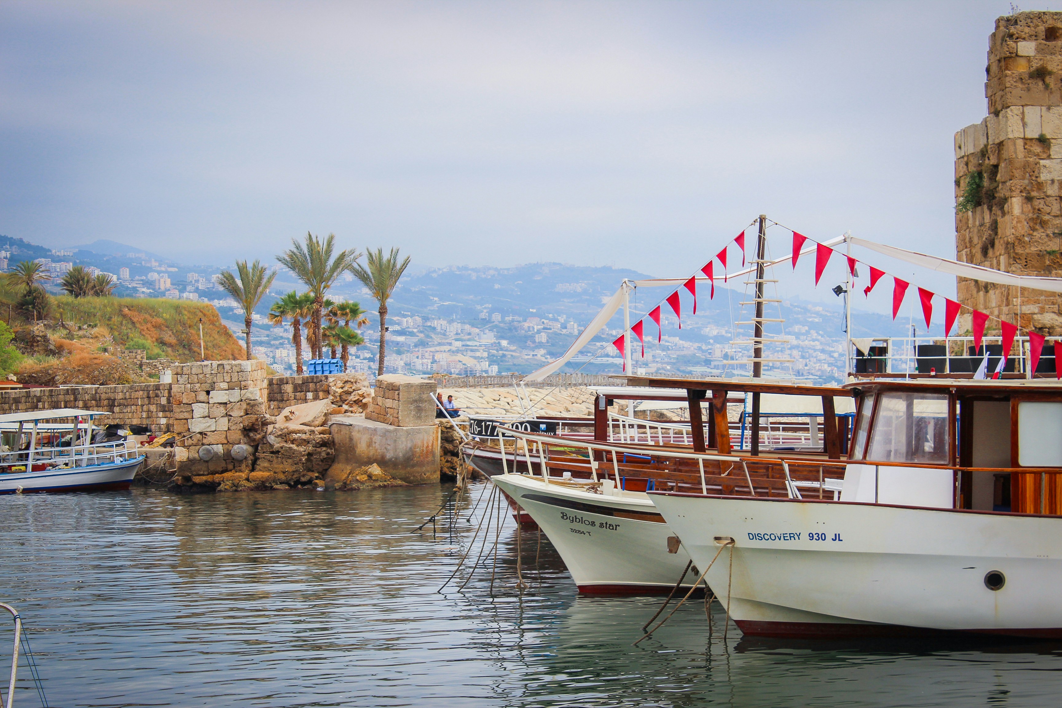Wooden boats docked in a calm harbor with distant mountains and palm trees under a cloudy sky.
