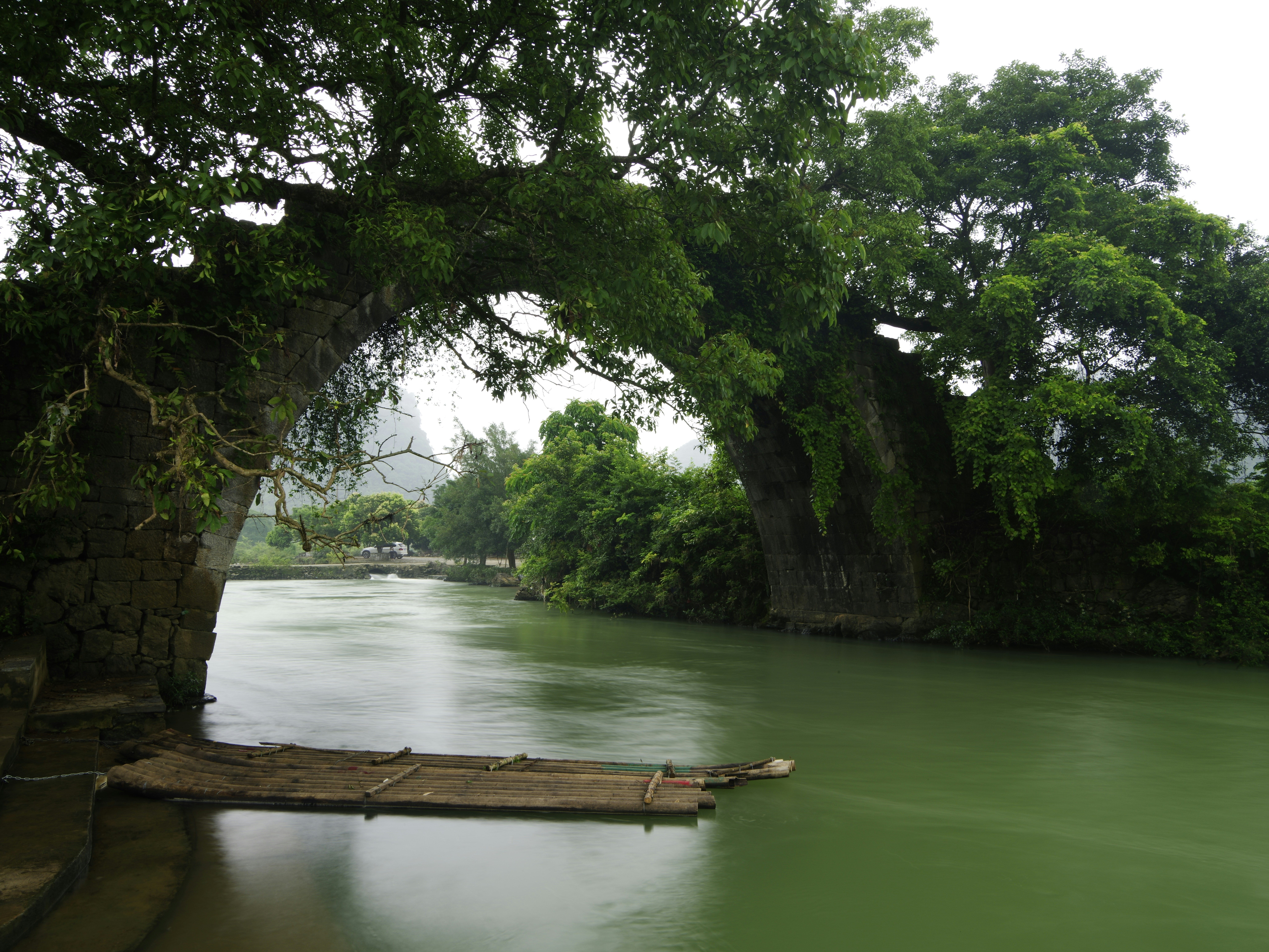 An ancient stone archway draped in lush greenery spans a tranquil river, reflecting the serene beauty of the surrounding landscape.