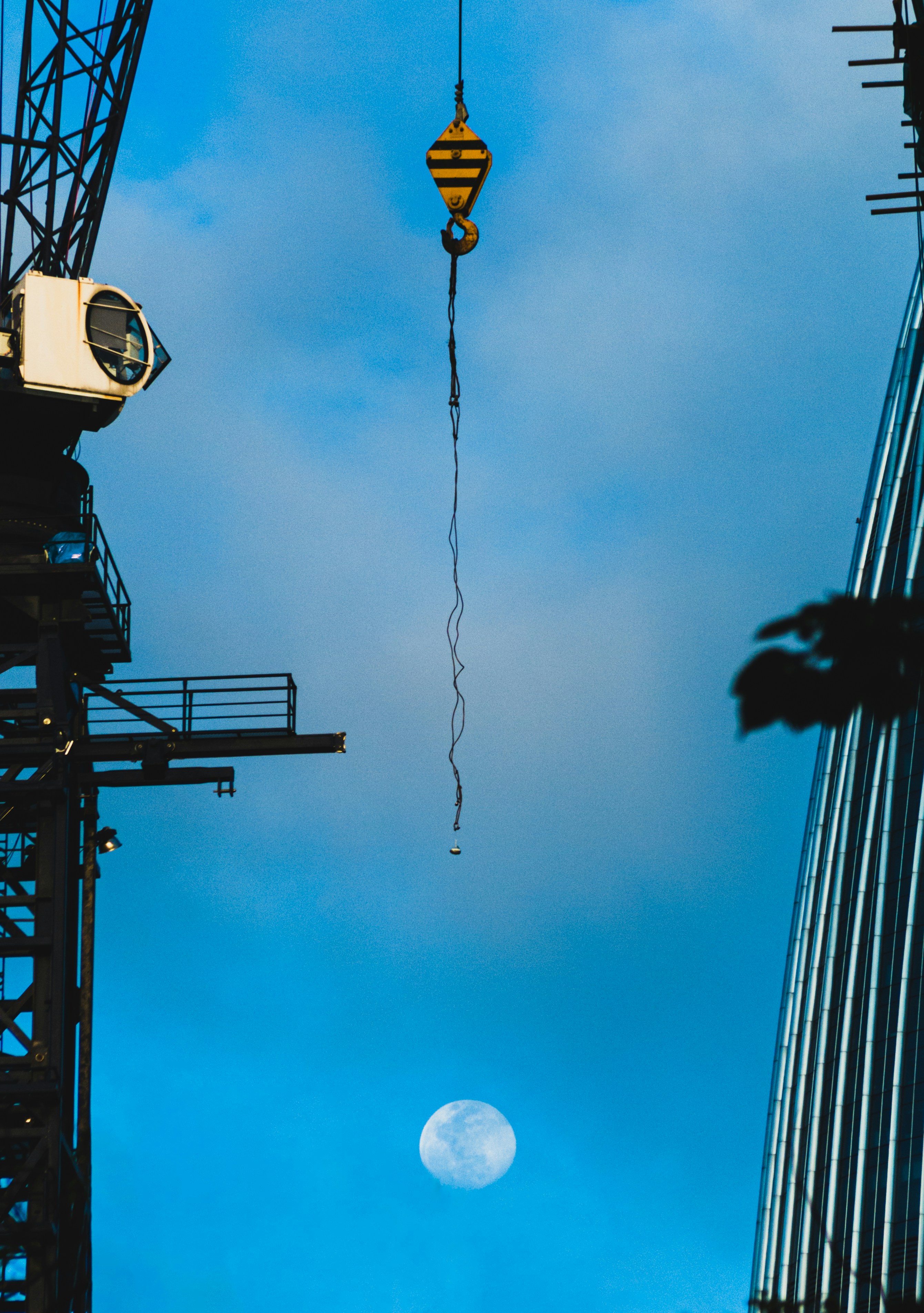 Crane hook descends from a construction tower into a clear blue sky, with the Moon visible low among the silhouettes of urban structures.