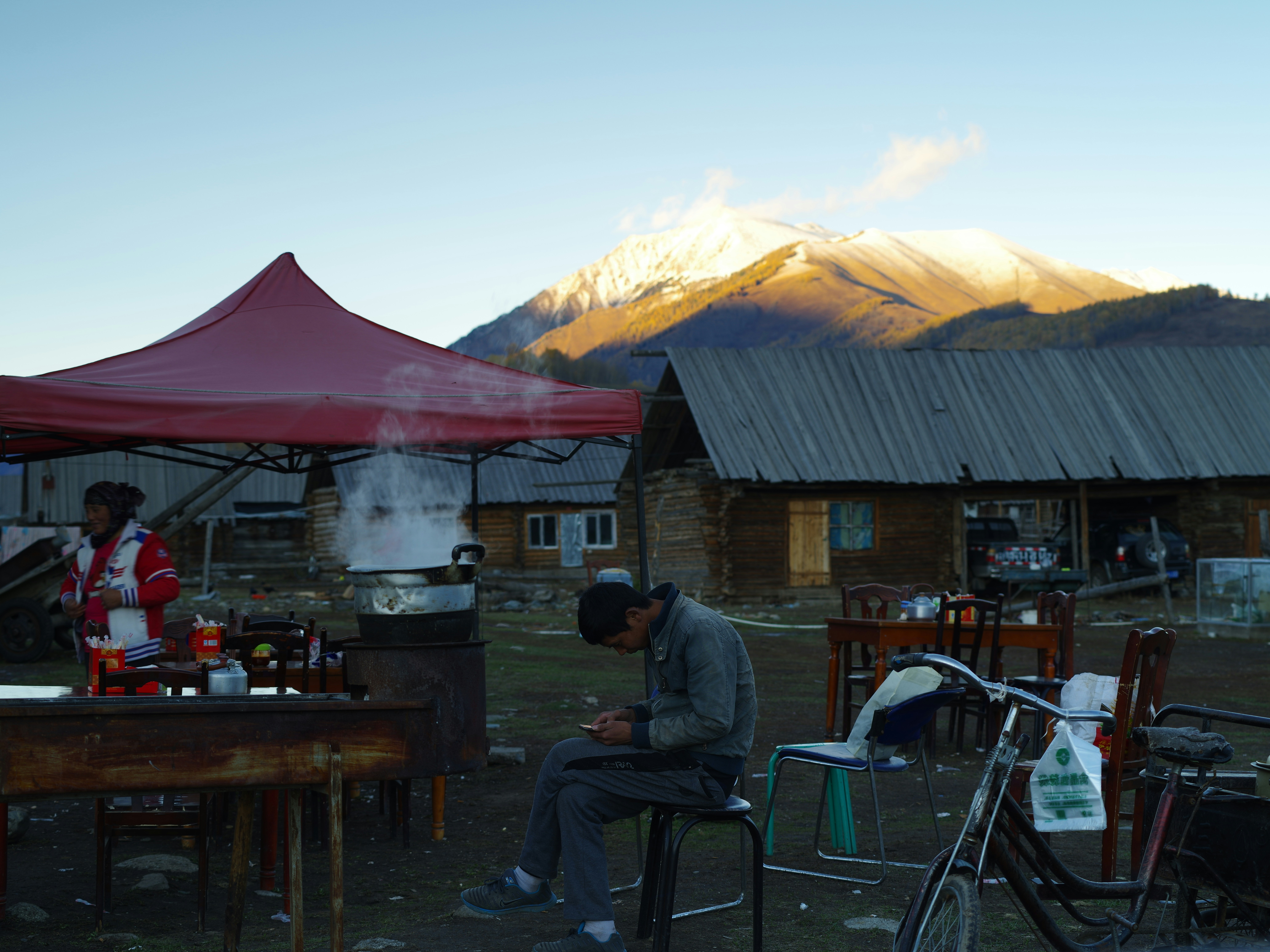 A lone figure sits contemplatively near a cooking station, with snow-capped mountains looming in the background. The scene captures a blend of daily life and nature's grandeur.