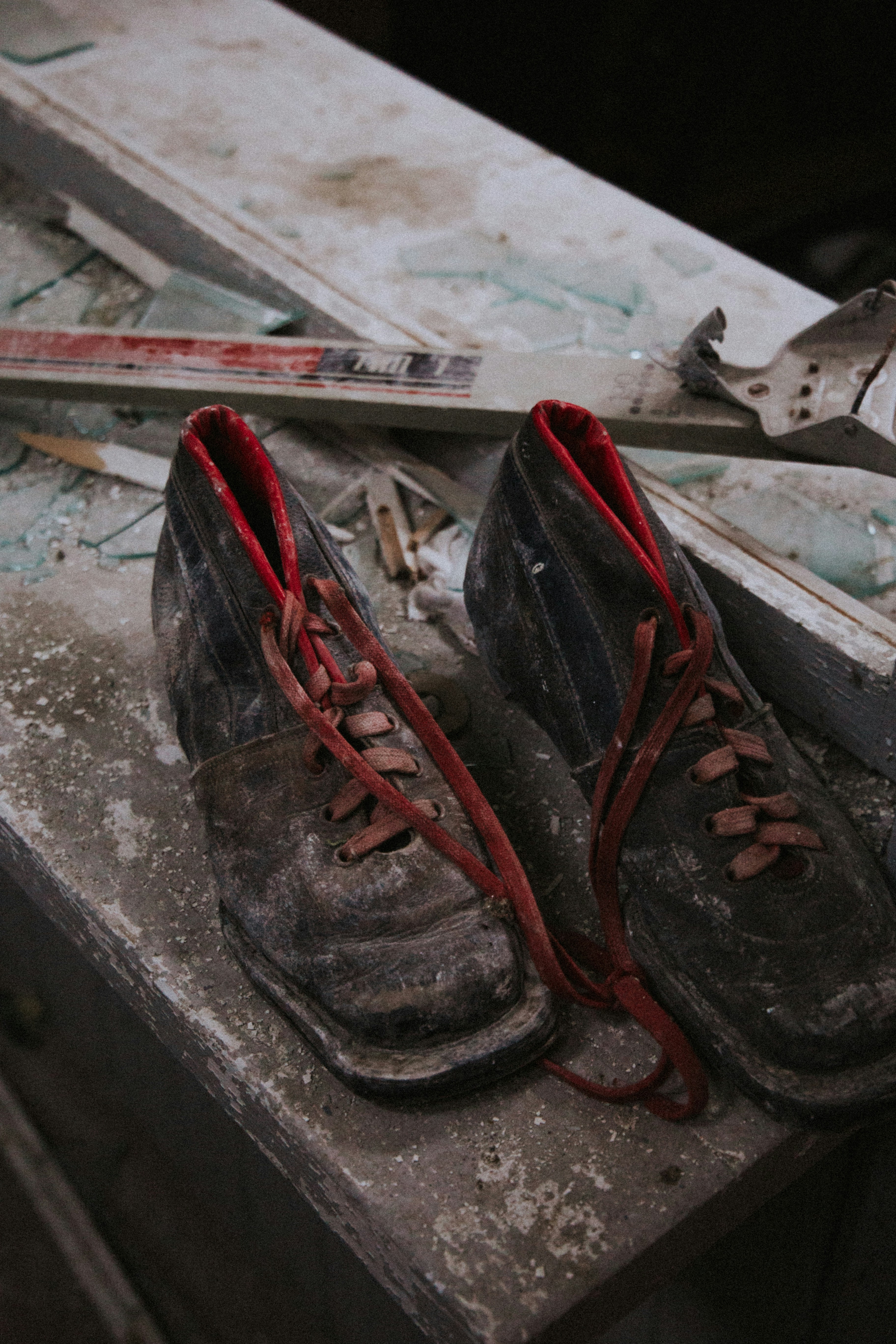 Worn vintage shoes with red laces resting on a dusty surface, surrounded by fragments of glass and debris.