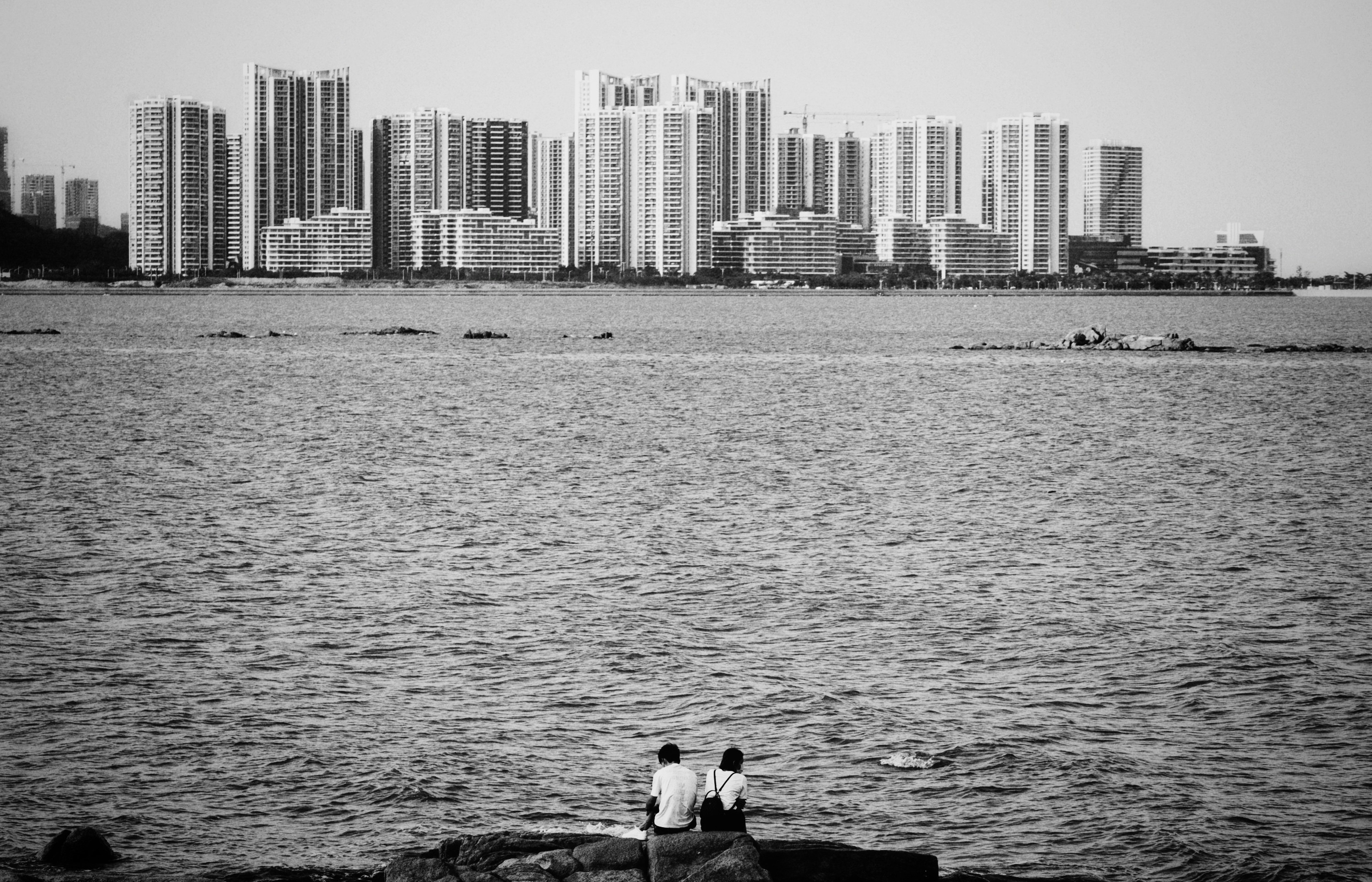Two people sitting on rocks by the sea with a city skyline in the distance.