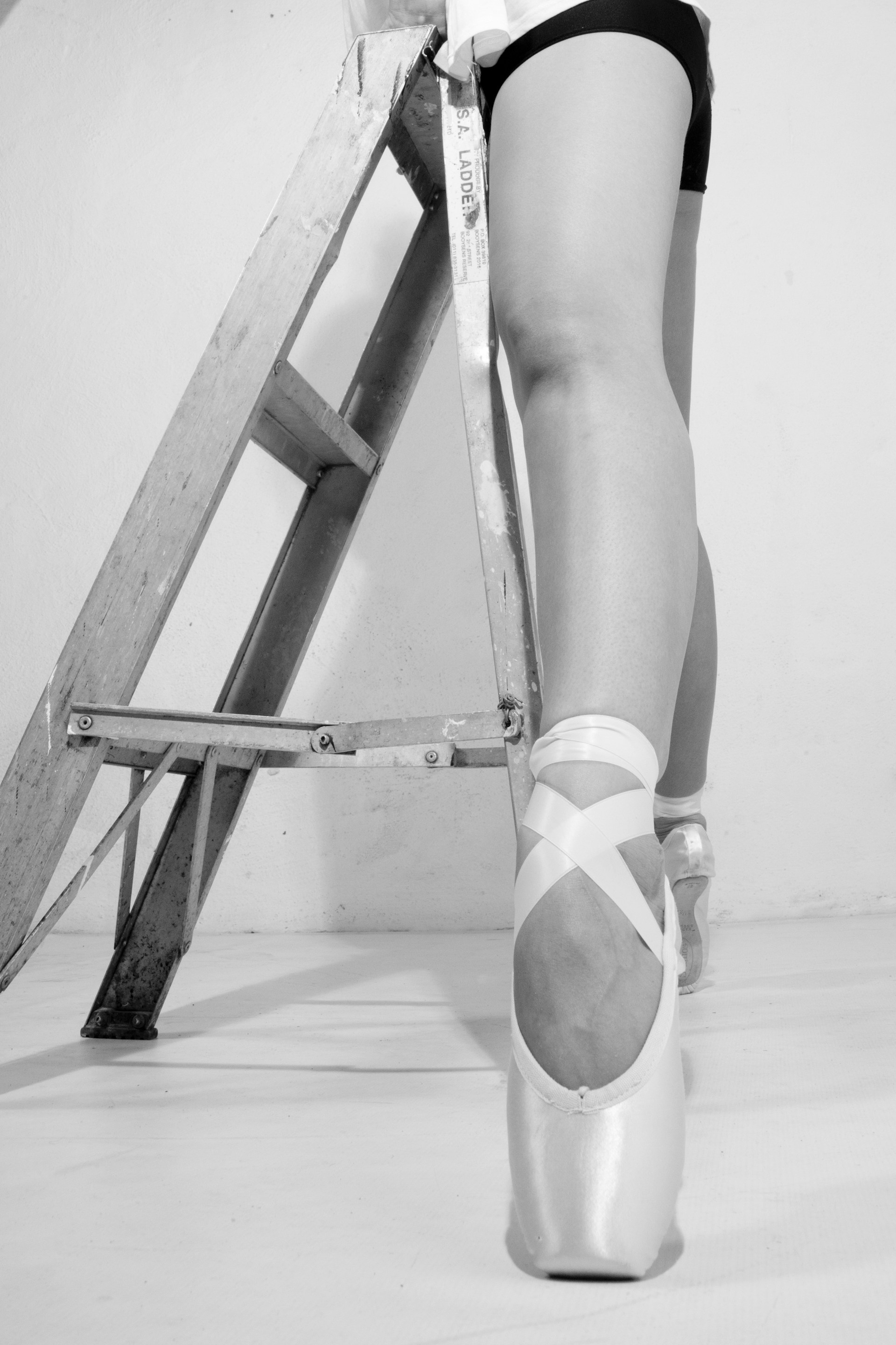 A dancer's leg in a pointe shoe steps near a weathered wooden ladder in a stark studio, captured in high-contrast black and white. The composition emphasizes form and balance against an austere backdrop.