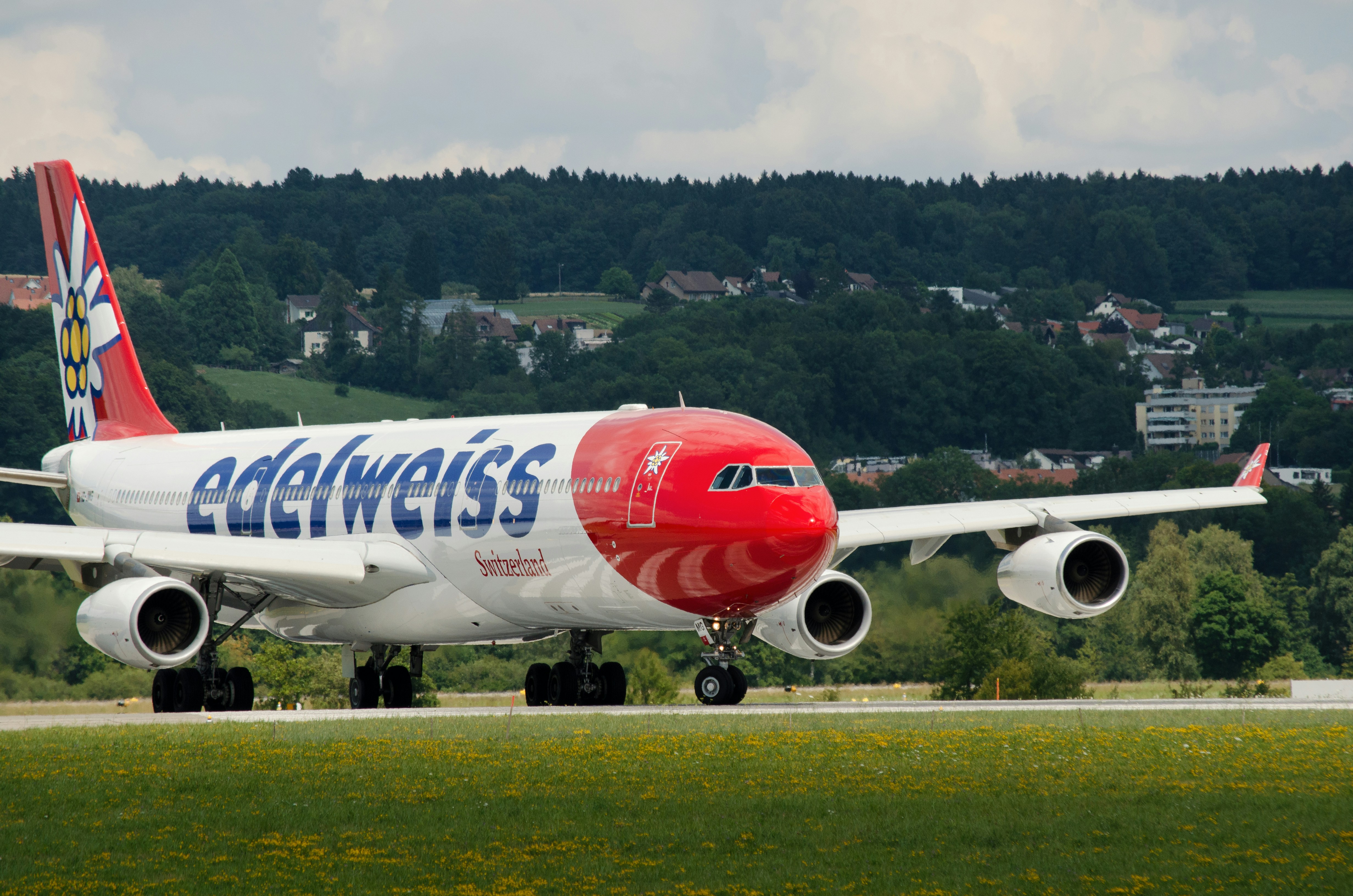 Airbus A340-300 with Edelweiss livery on taxiway at Zurich airport, framed by green hills and cloudy sky.