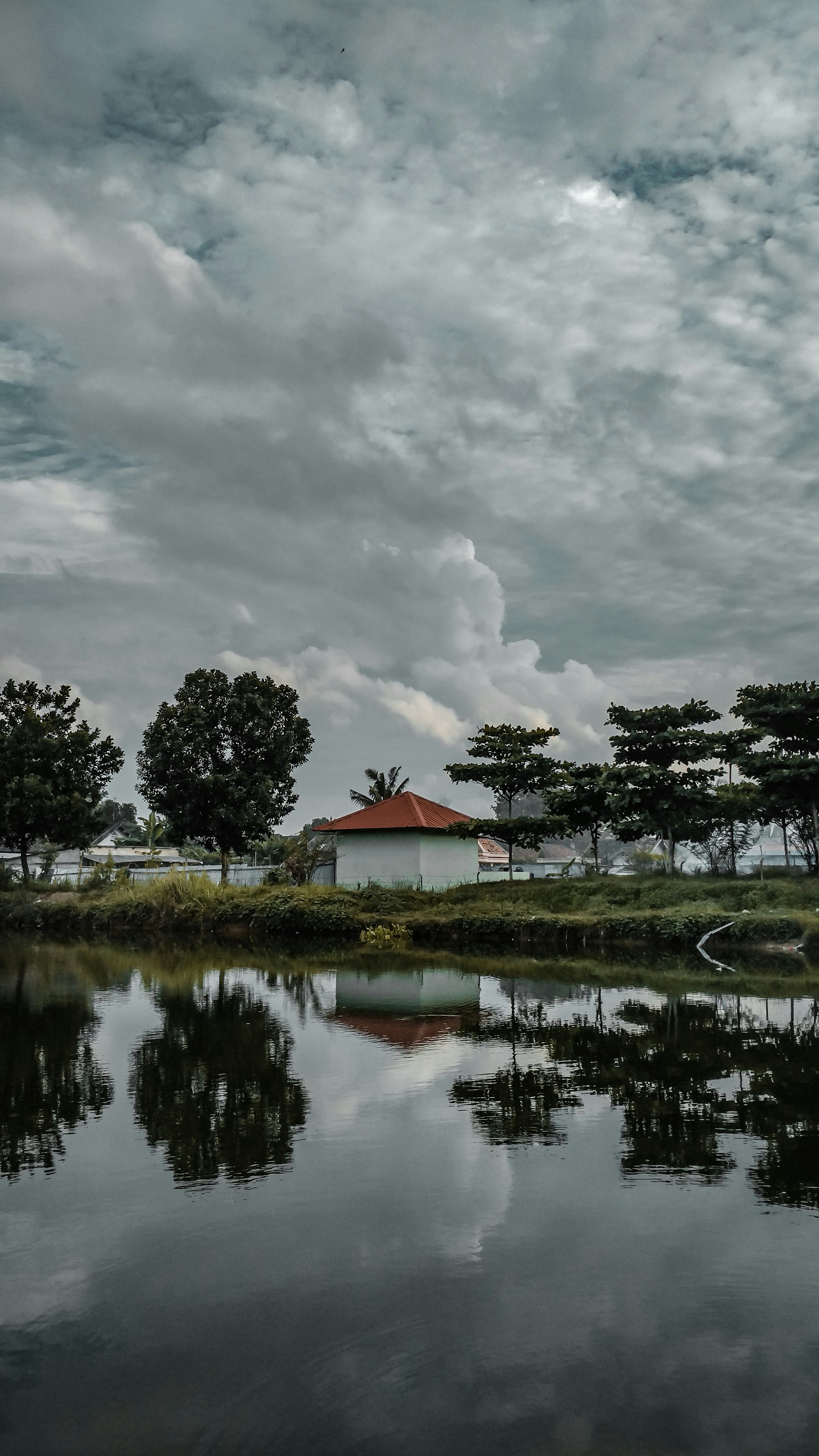 A tranquil lakeside scene featuring a white house with a red roof, surrounded by lush trees and mirrored in calm waters. The overcast sky adds a serene ambiance.
