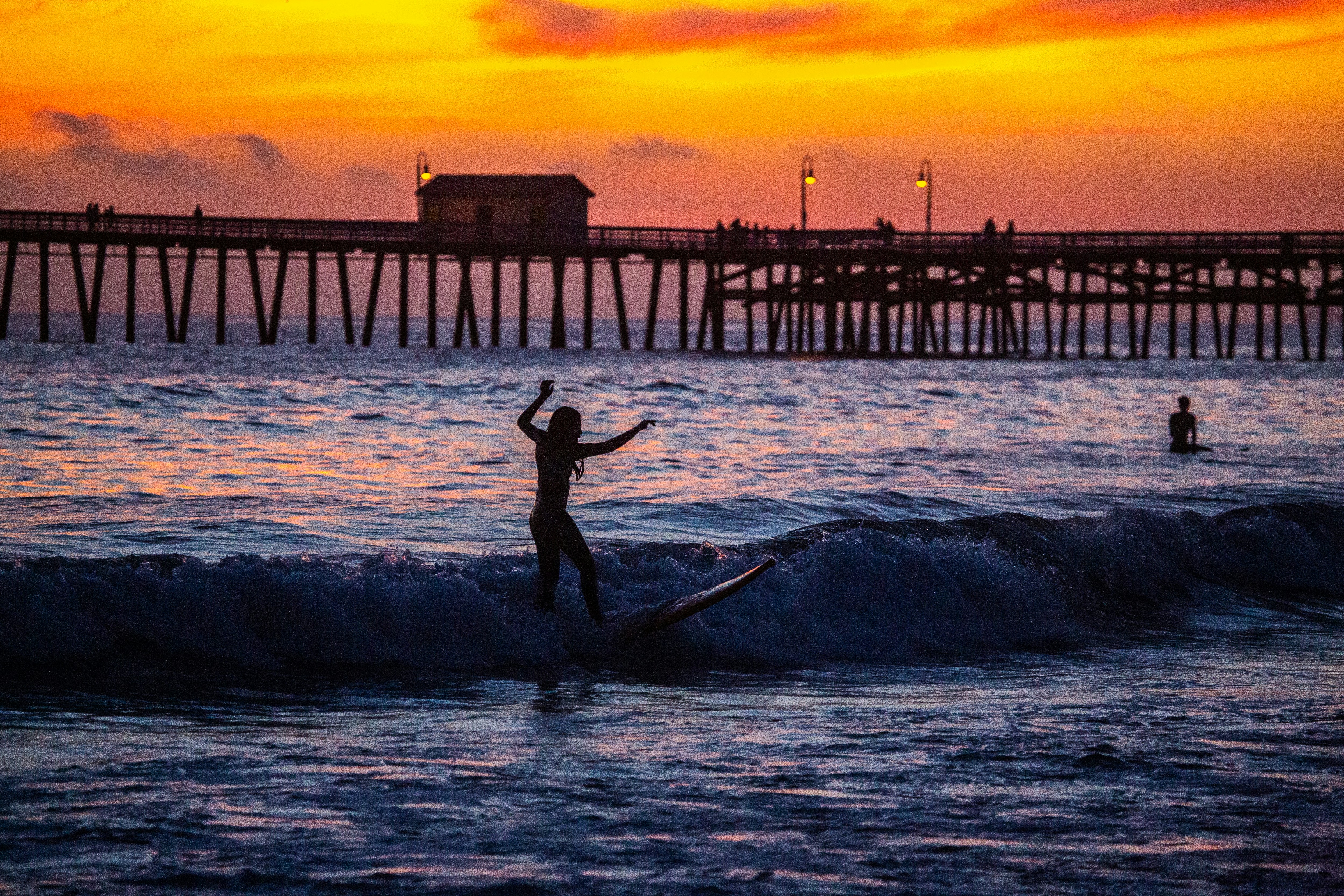 Surfer riding a wave at sunset near a lit pier with vibrant orange and purple sky.