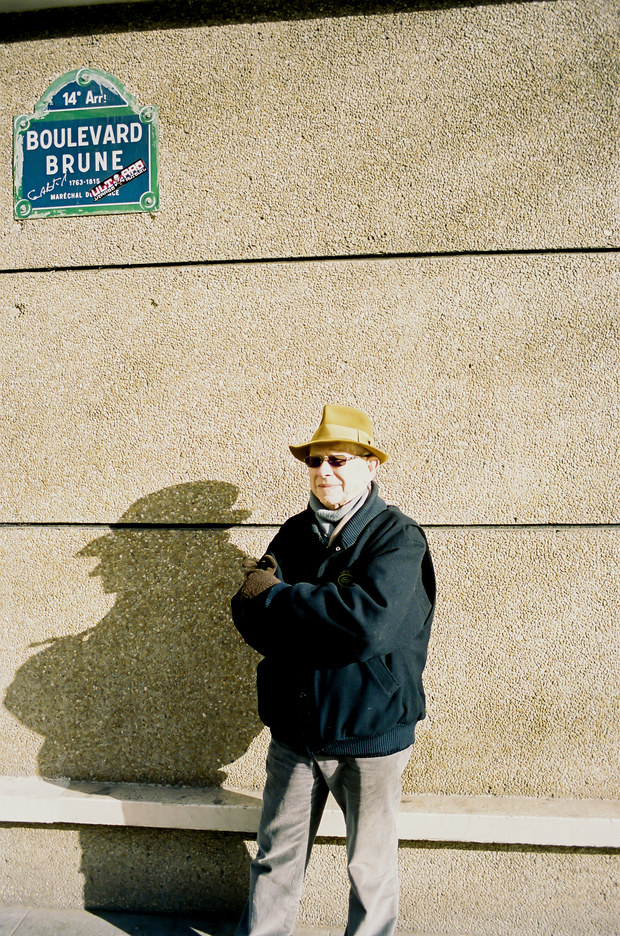 A man in a tan fedora and dark jacket stands against a textured beige wall beneath a Boulevard Brune street sign in Paris.