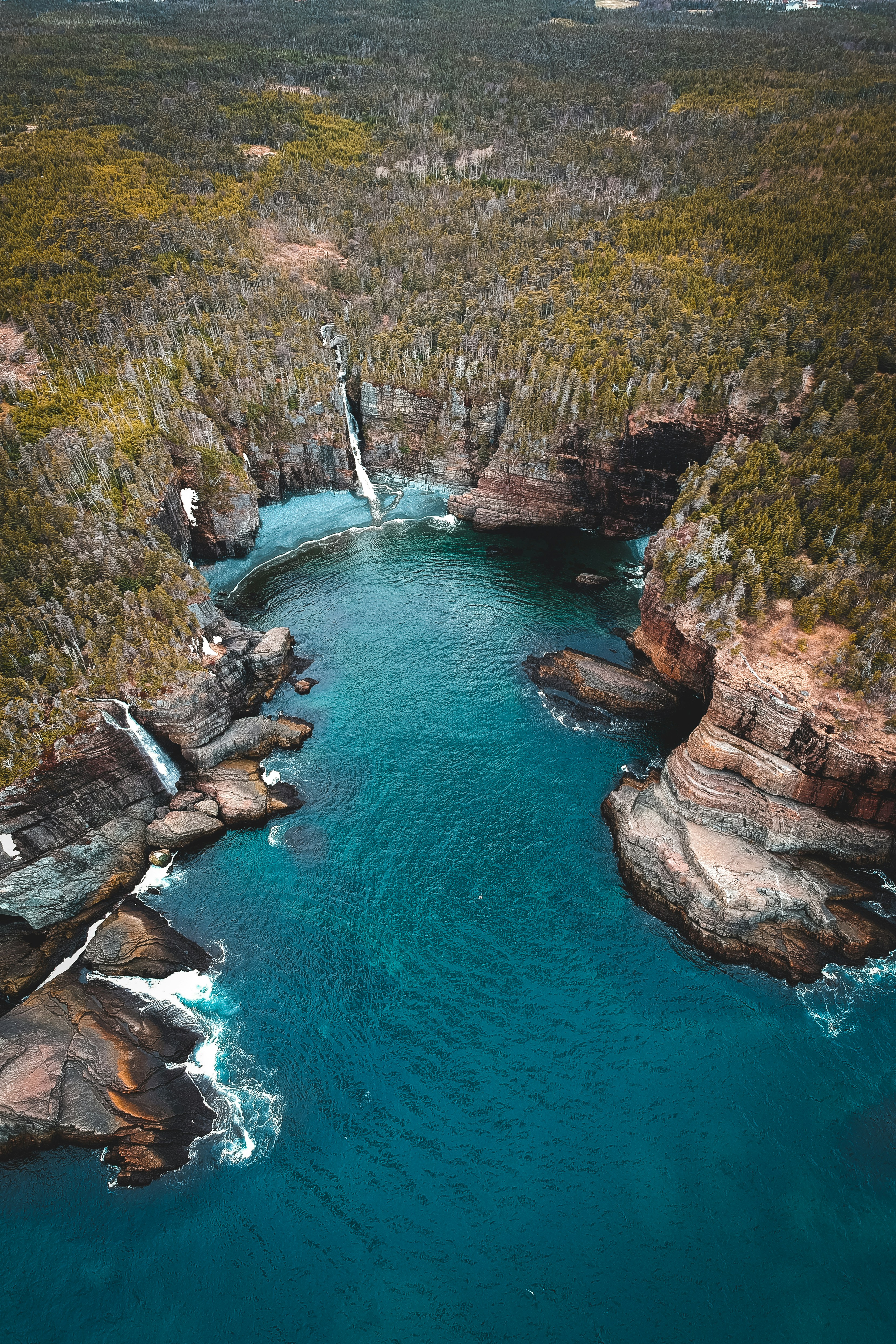 Aerial view of a secluded cove surrounded by lush greenery and rocky cliffs, featuring cascading waterfalls into the turquoise waters below.