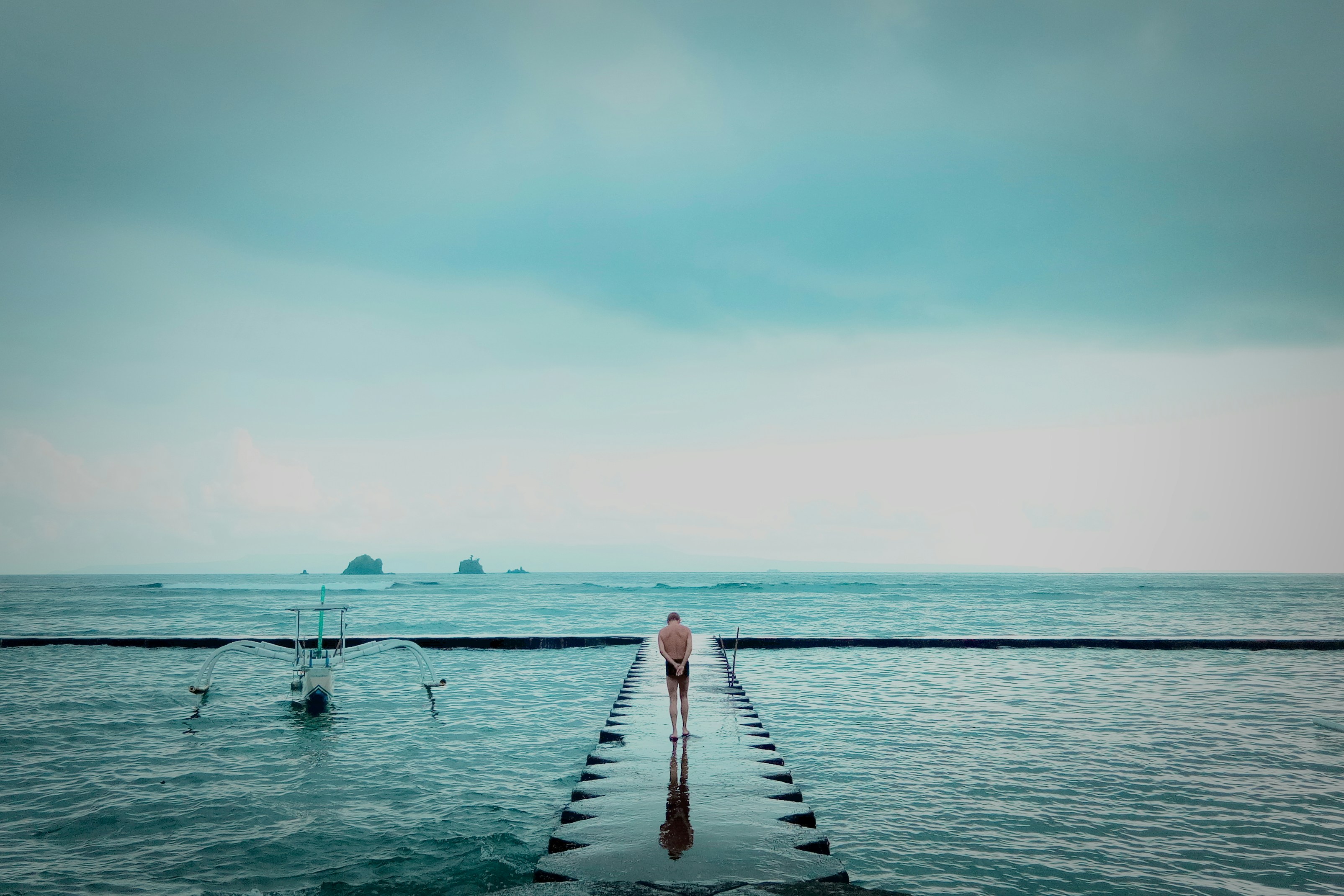 A solitary figure stands at the end of a long pier extending into a calm sea beneath a moody blue sky.