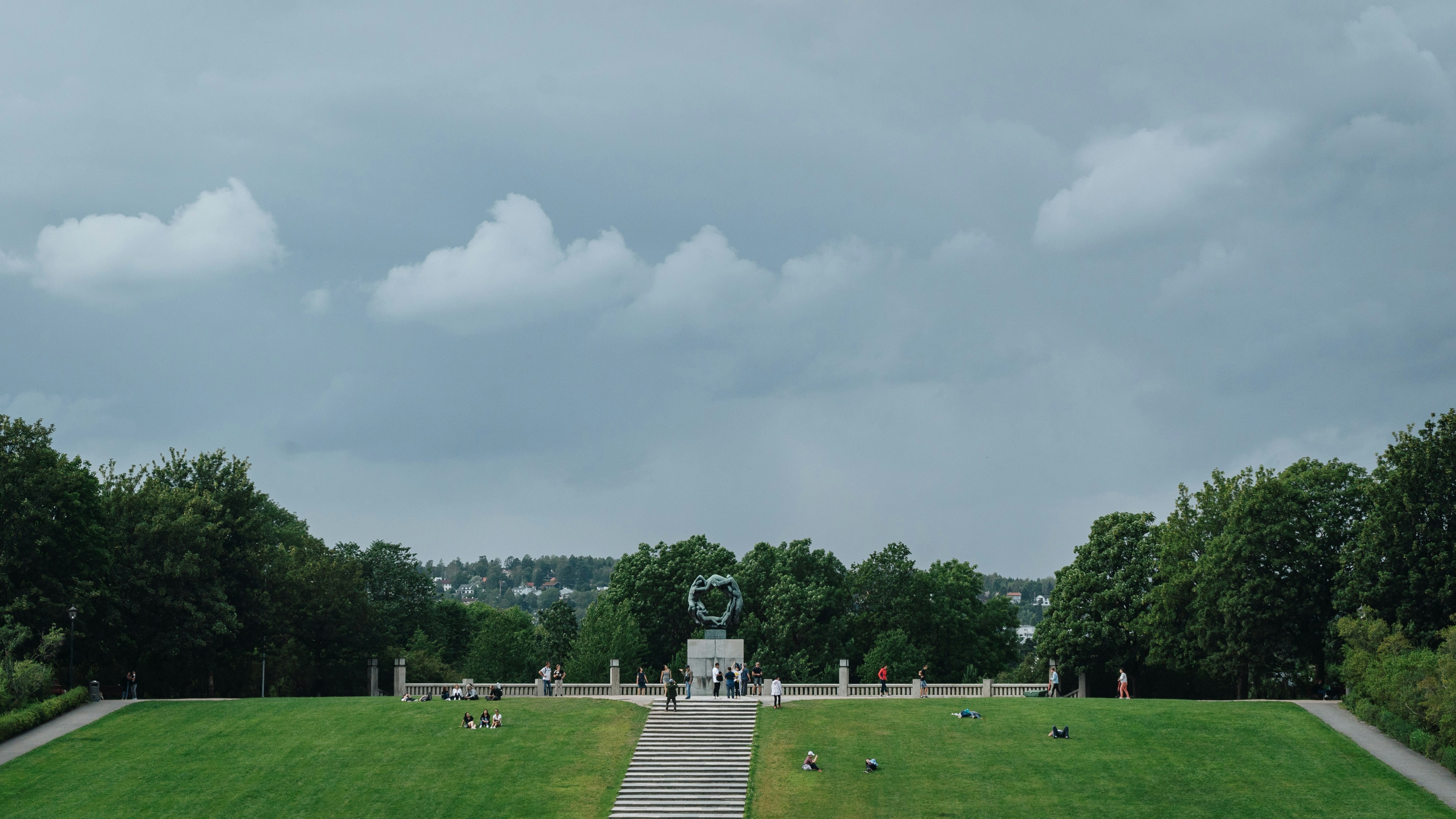Wide view of a park with visitors scattered on the grass, framed by lush trees under a dramatic sky. A monument stands at the center, inviting contemplation.
