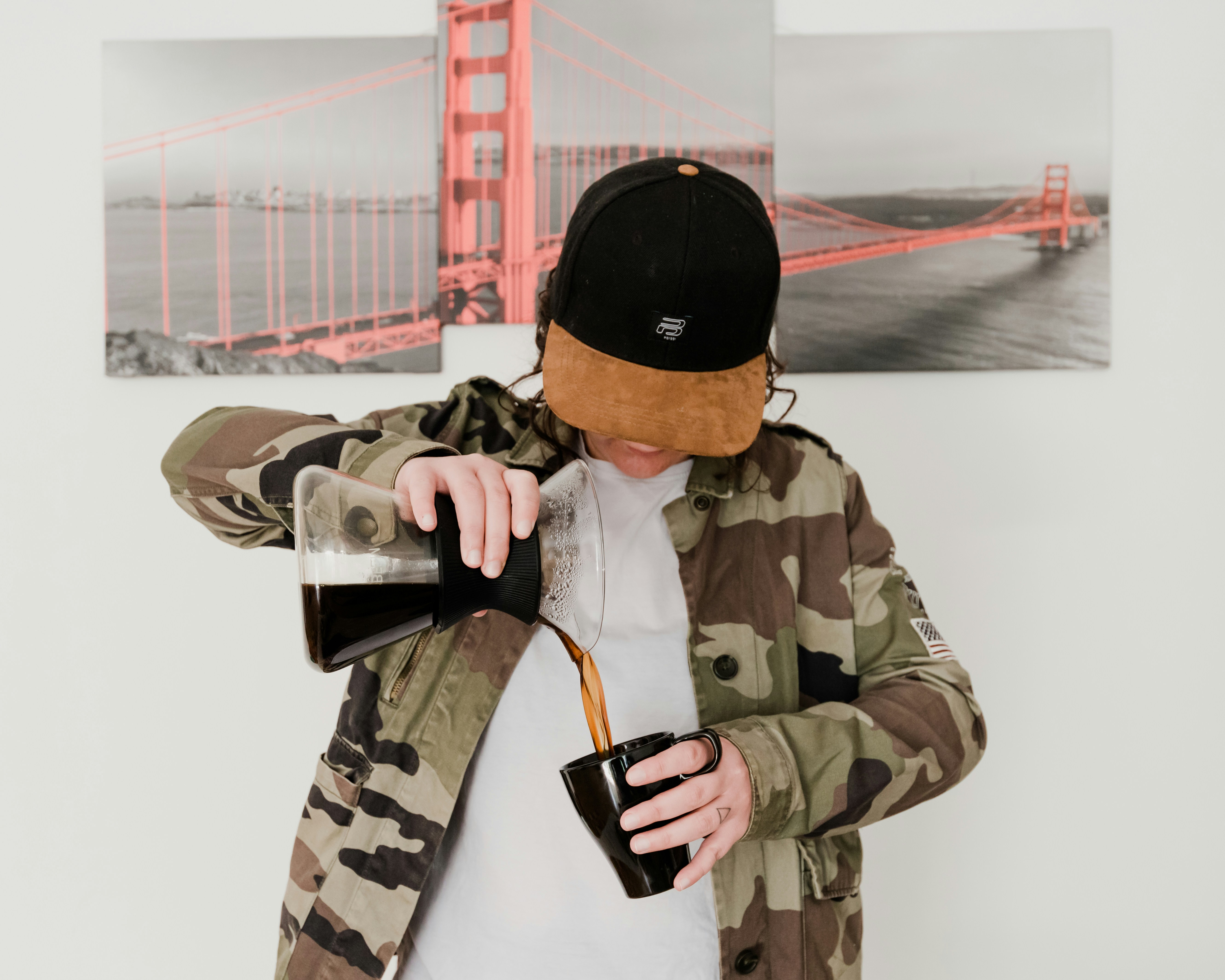 Person in camouflage jacket pouring coffee from a glass carafe into a black cup, with a backdrop of the Golden Gate Bridge in vibrant hues.