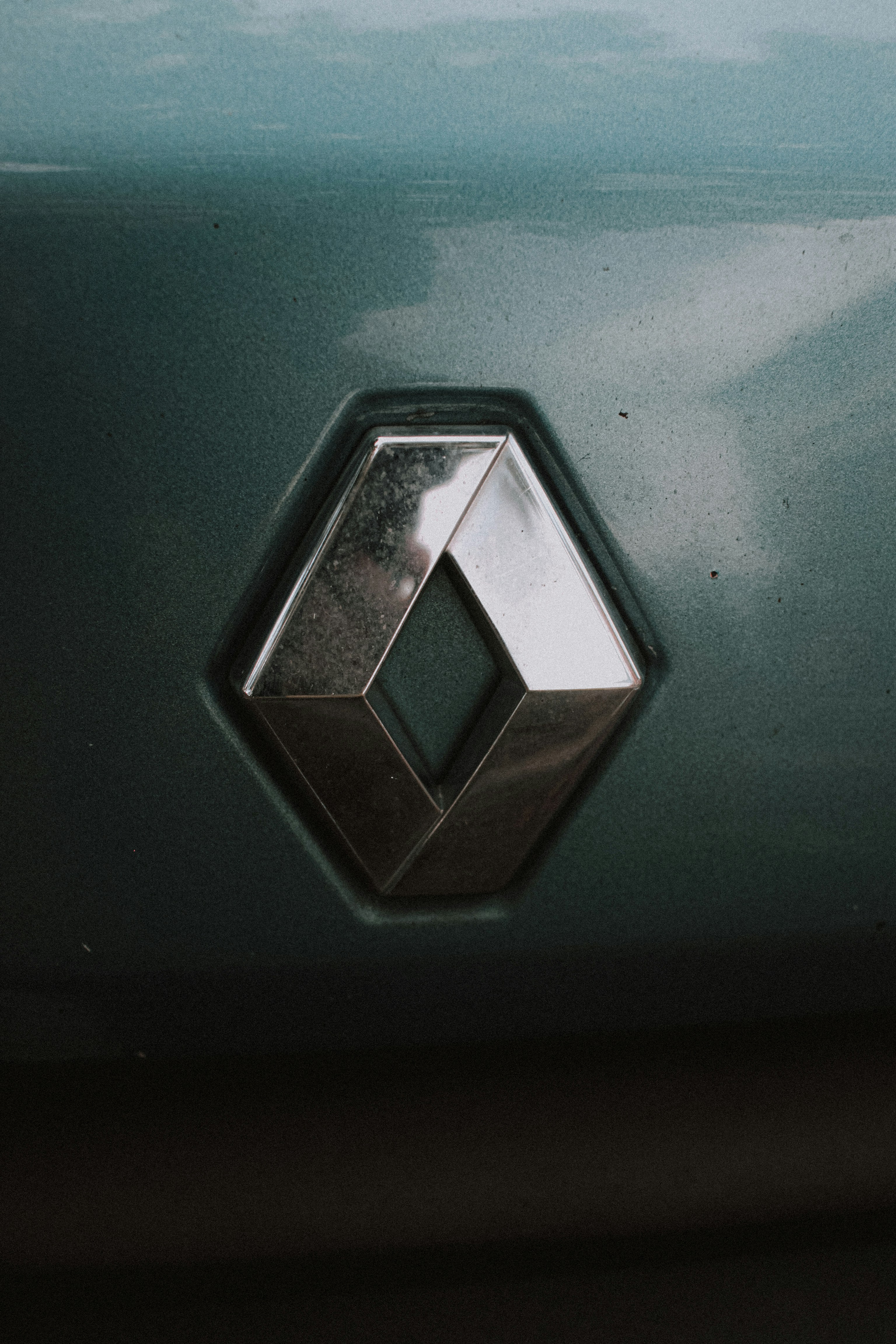 Close-up of the Renault logo reflecting light against a textured teal surface. The emblem showcases the brand's iconic diamond shape.