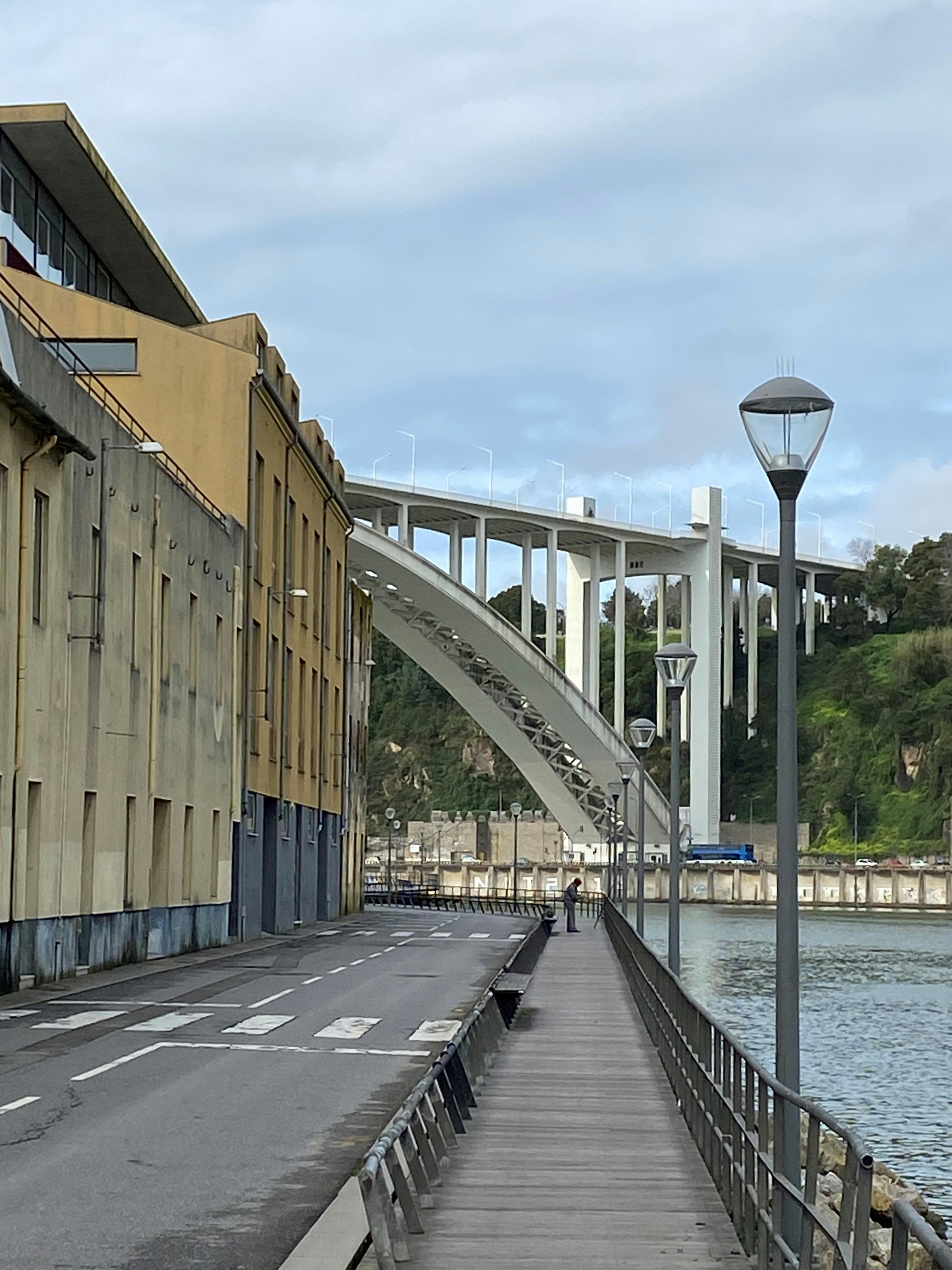 A riverside pathway lined with street lamps, leading towards a modern bridge that arches gracefully over the water, complemented by a backdrop of lush greenery.