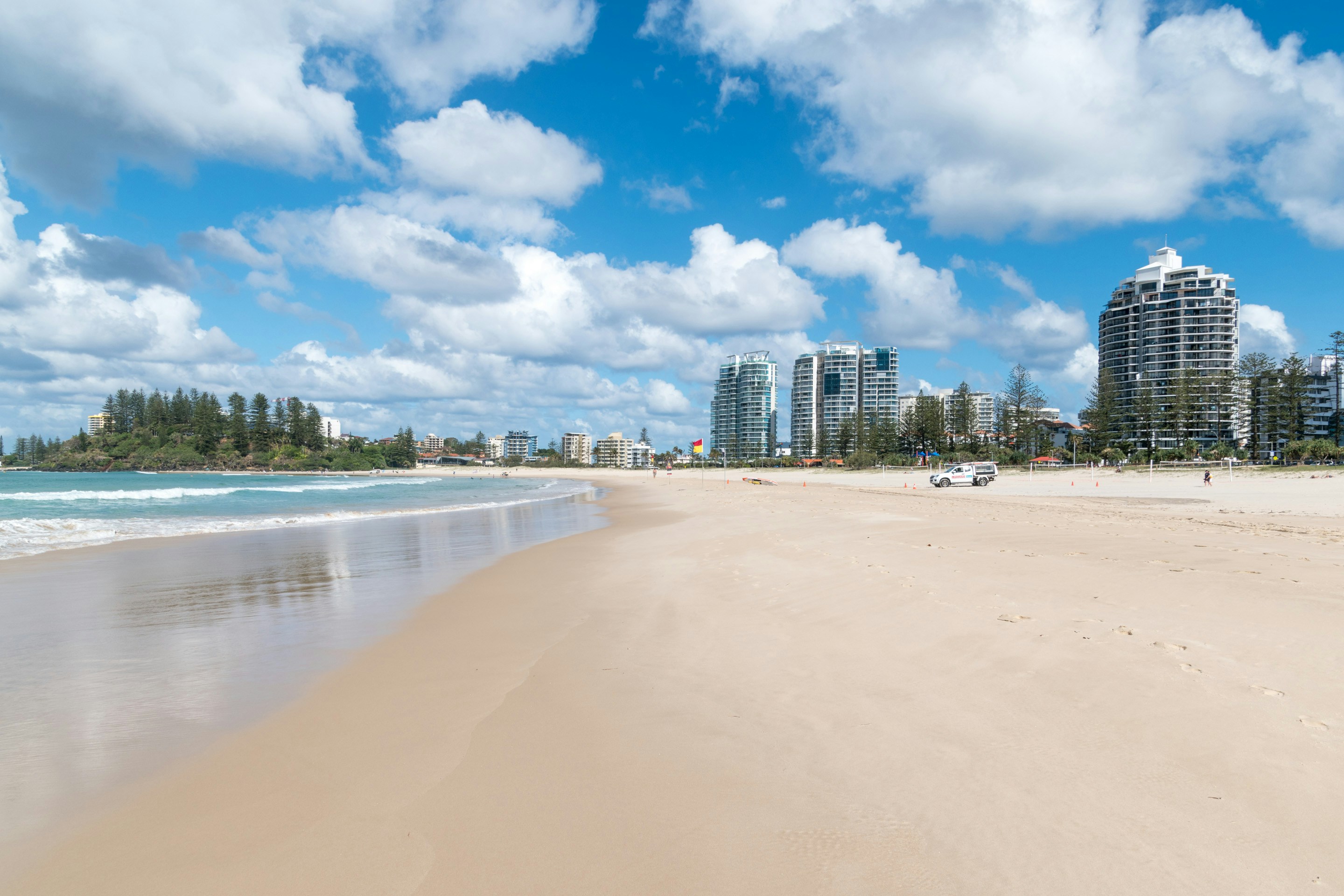 Expansive beach scene with gentle waves lapping at the shore and modern buildings lining the coastline under a partly cloudy sky.