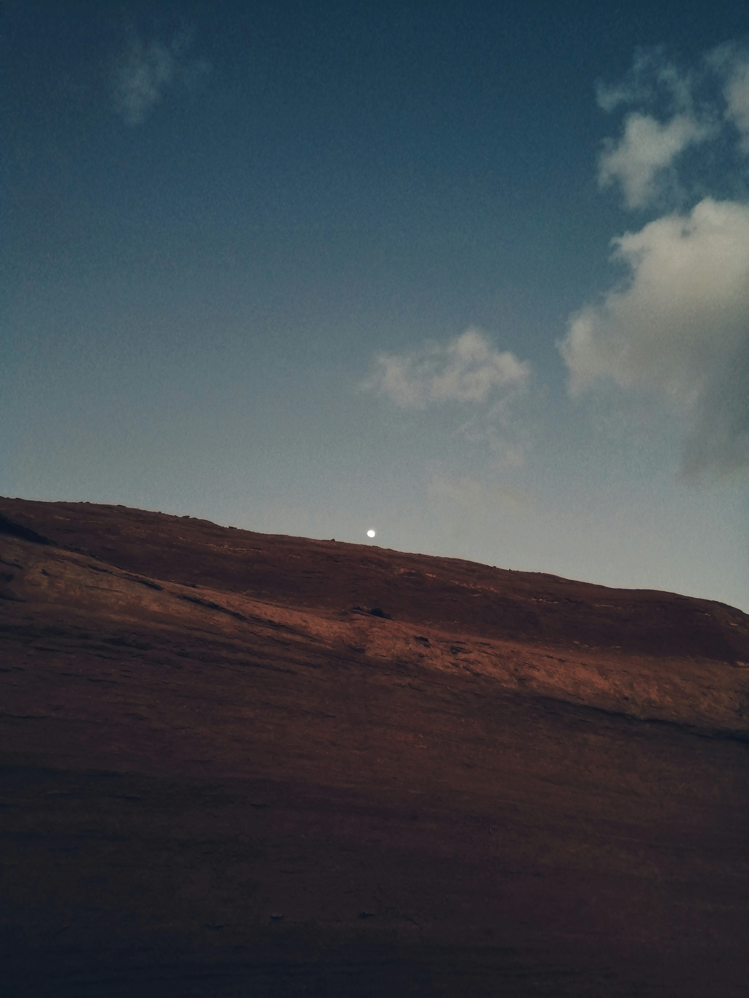 The moon rises gently over a rugged landscape, casting a soft glow against the twilight sky. Clouds drift lazily, enhancing the serene atmosphere.