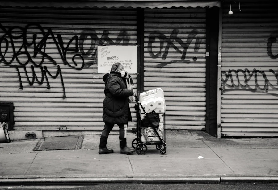 A masked pedestrian in a heavy coat pushes a small cart loaded with supplies along a graffiti-covered, closed storefront. The scene is rendered in black and white with an urban, documentary feel.