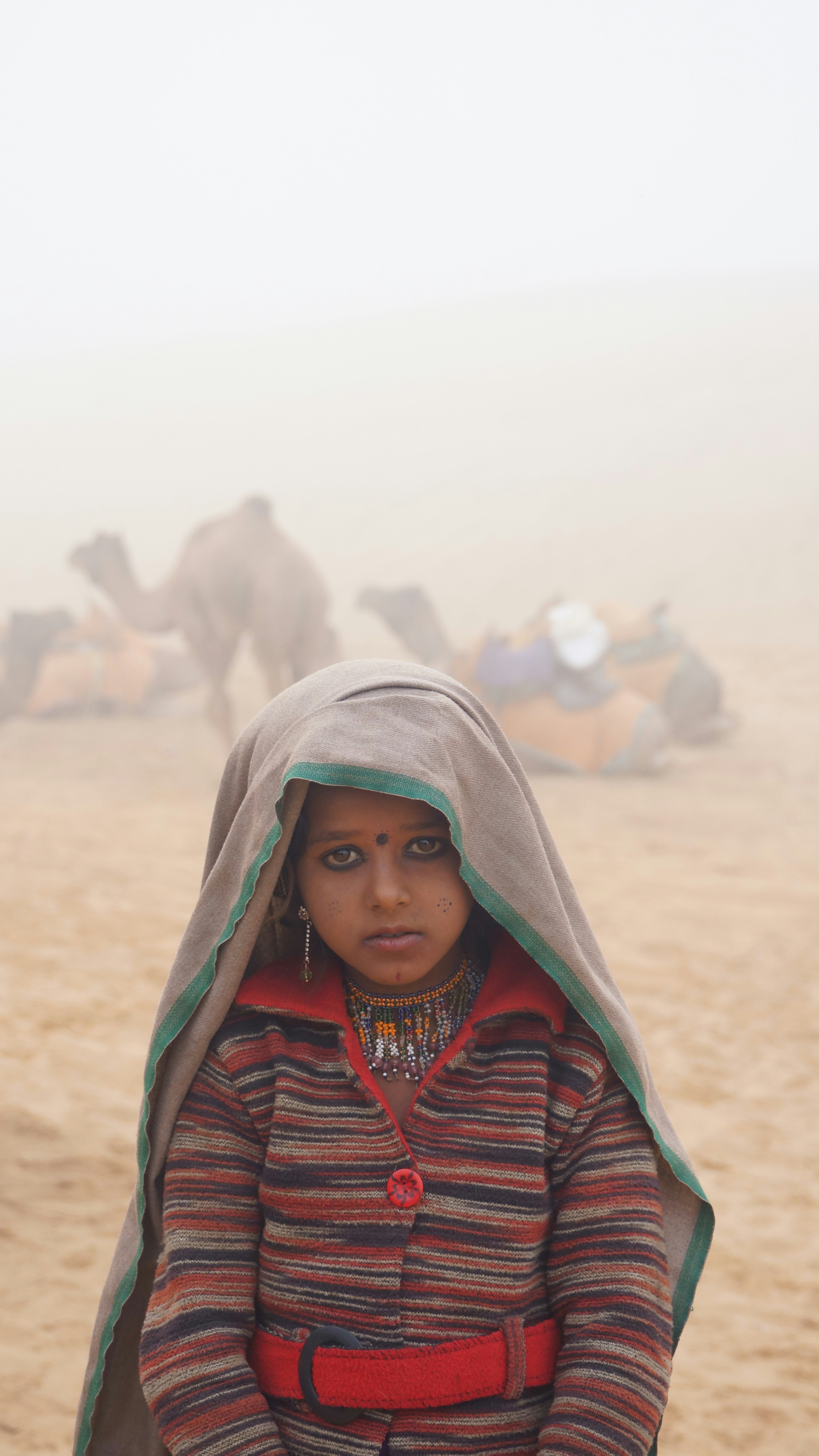 A young child draped in a gray shawl stands against a foggy desert backdrop, with camels resting in the distance. The image captures a moment of quiet resilience.