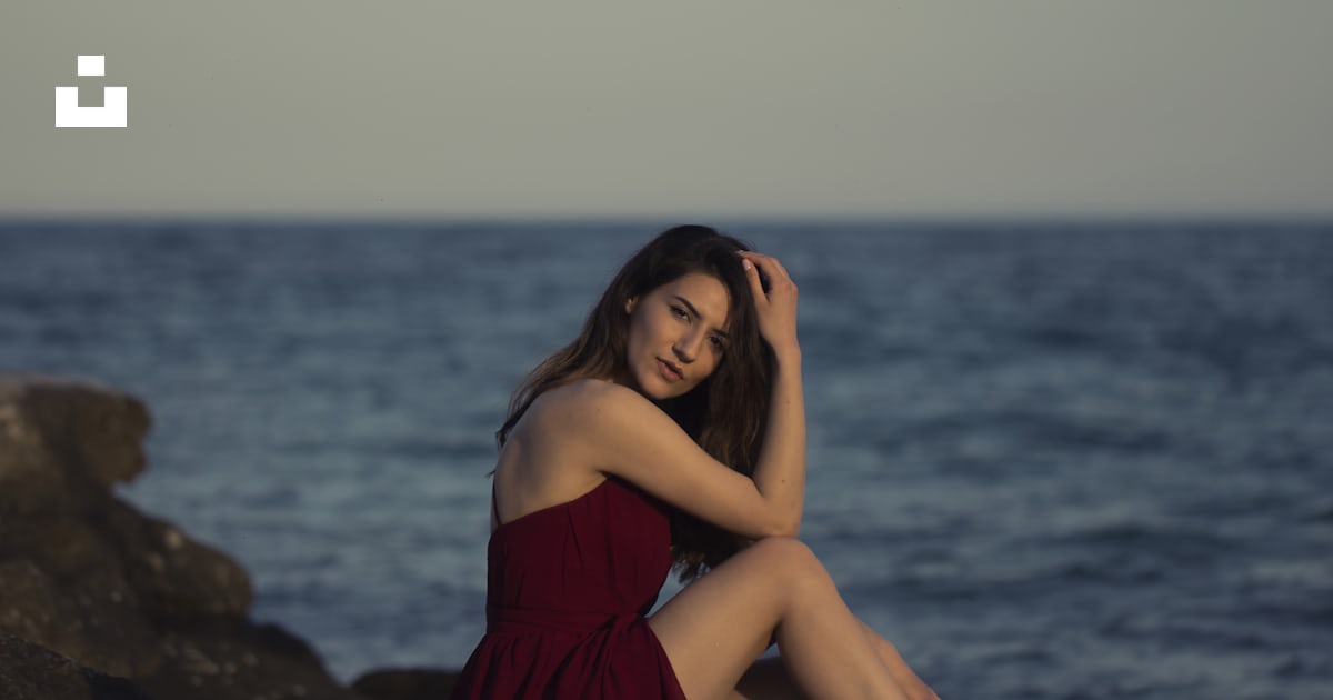 Woman In Red Dress Sitting On Rock Near Sea During Daytime Photo Free