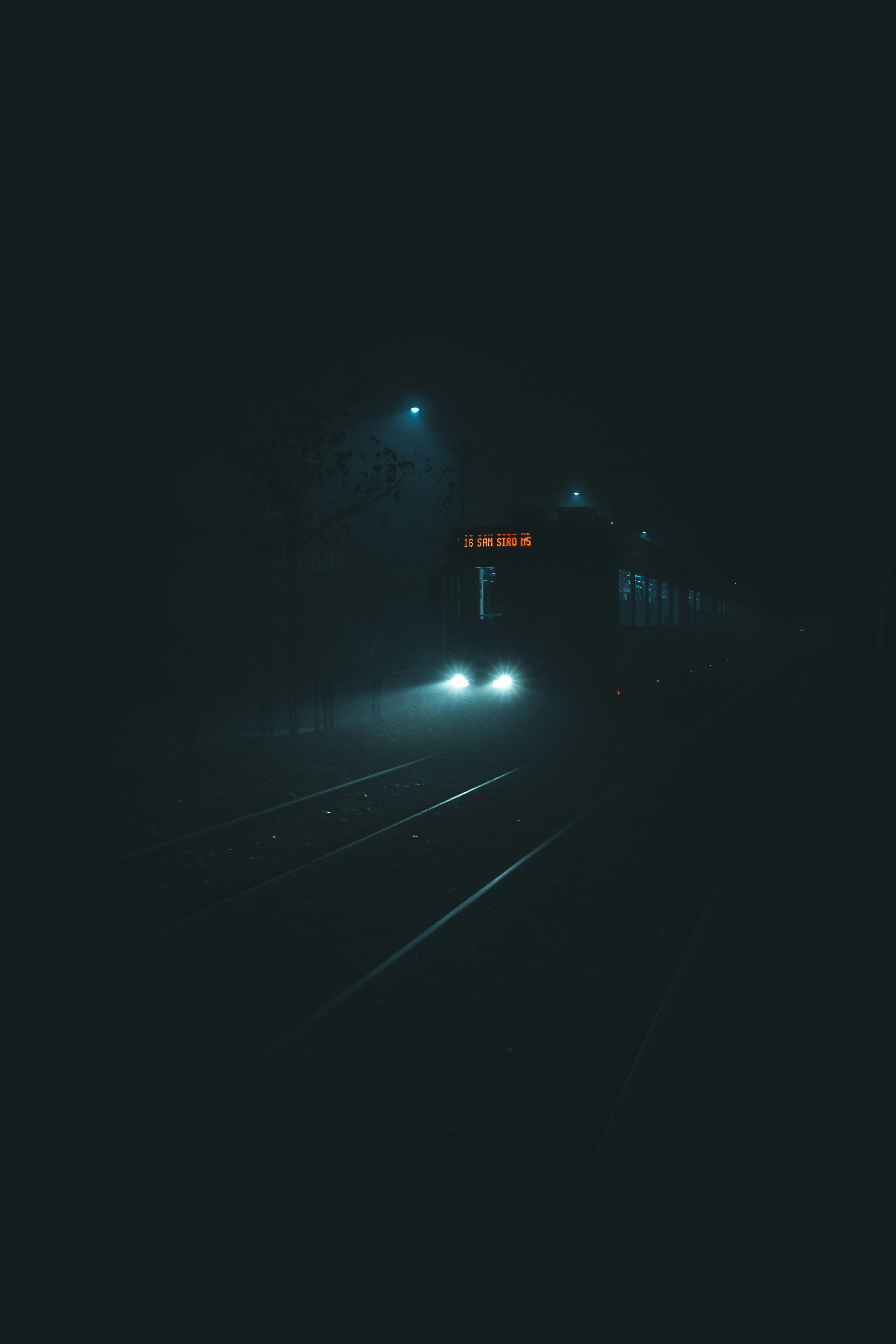 A train emerges from the fog, its headlights piercing the darkness, with illuminated signage hinting at its destination.