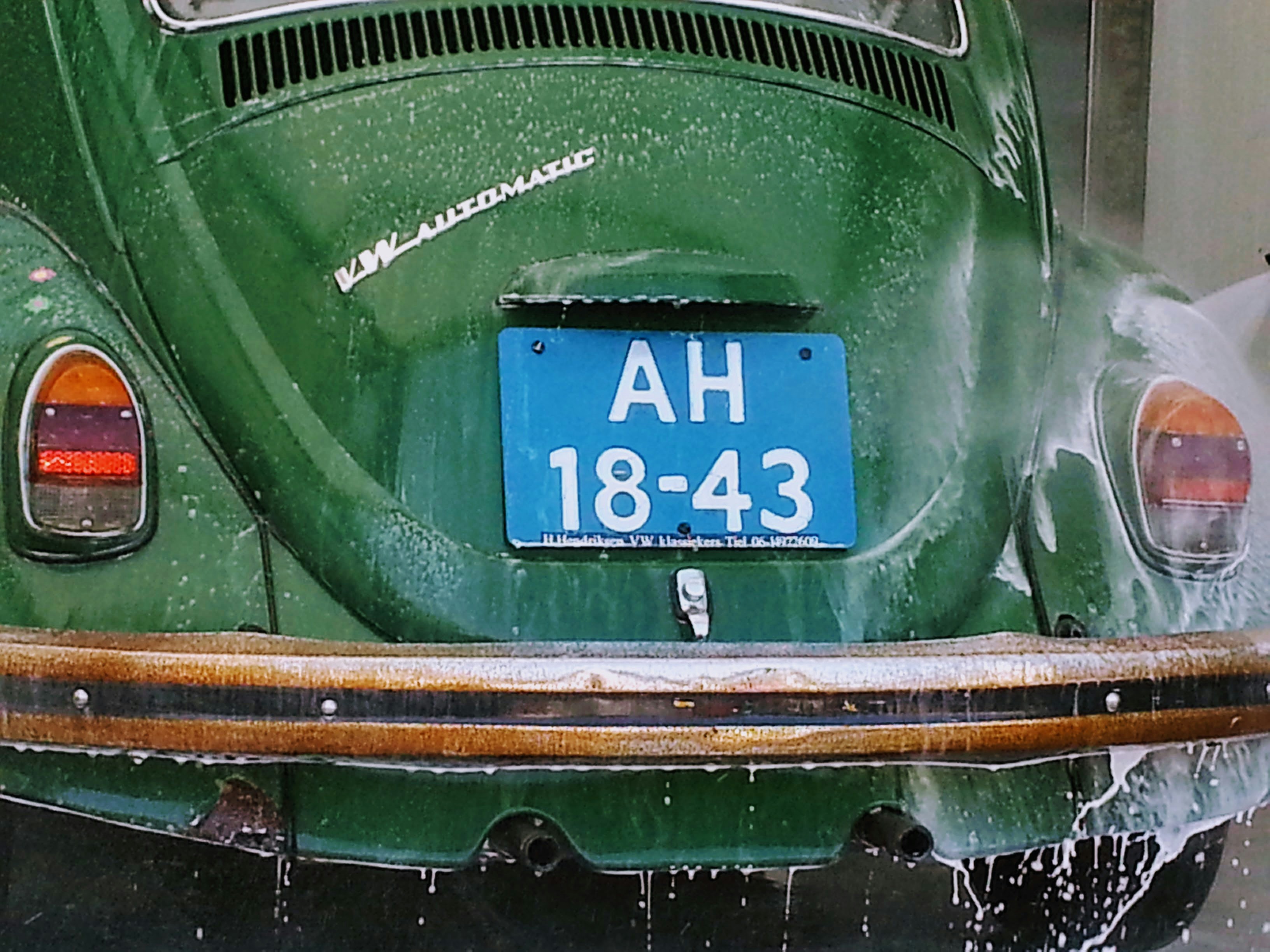 Classic green Volkswagen Beetle being washed, with soap suds cascading down its rear. The vibrant blue license plate stands out against the shiny finish.
