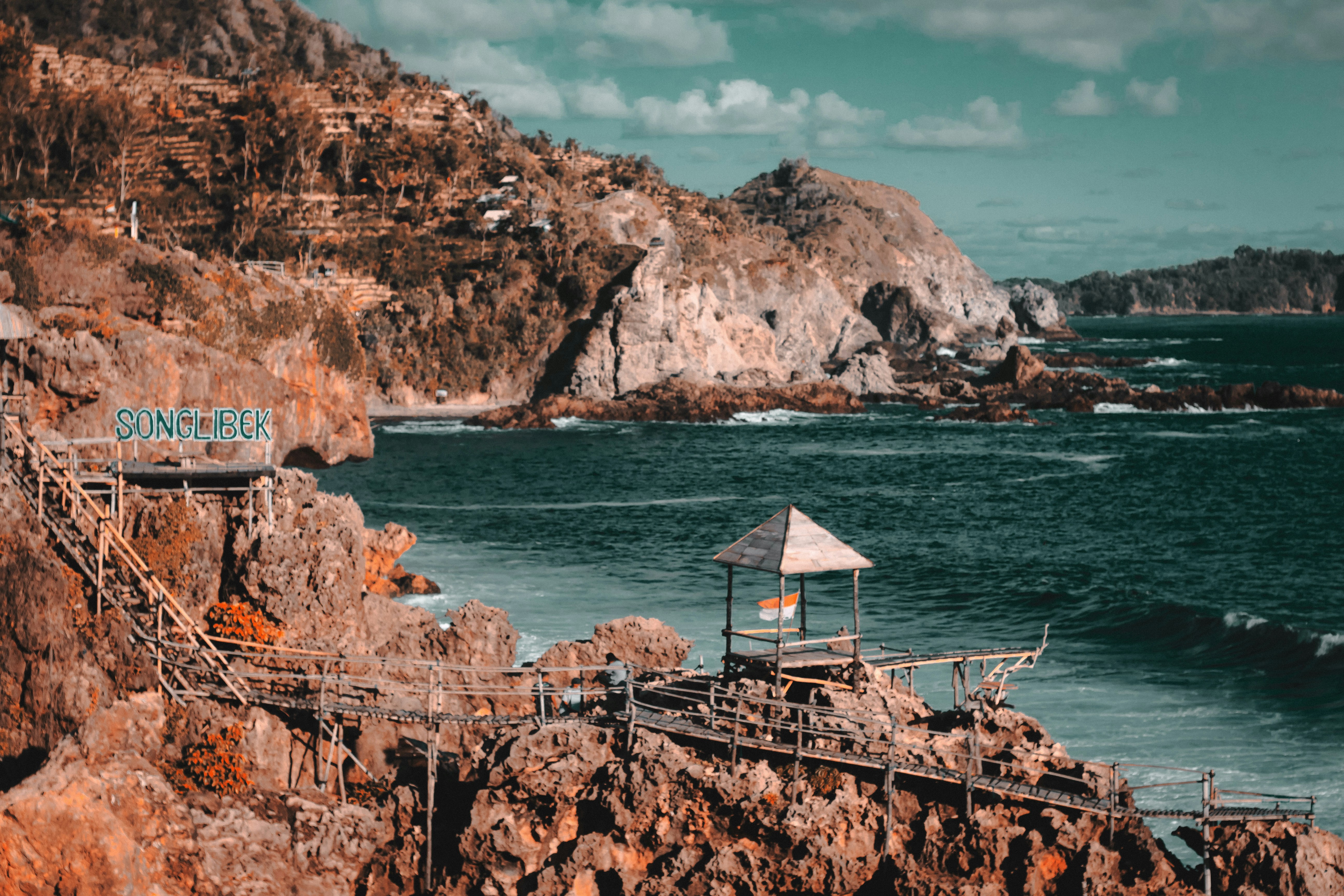 Rustic wooden pavilion perched on rocky coastline, overlooking the ocean with vibrant blue waters and distant cliffs. The scene conveys a tranquil escape into nature.