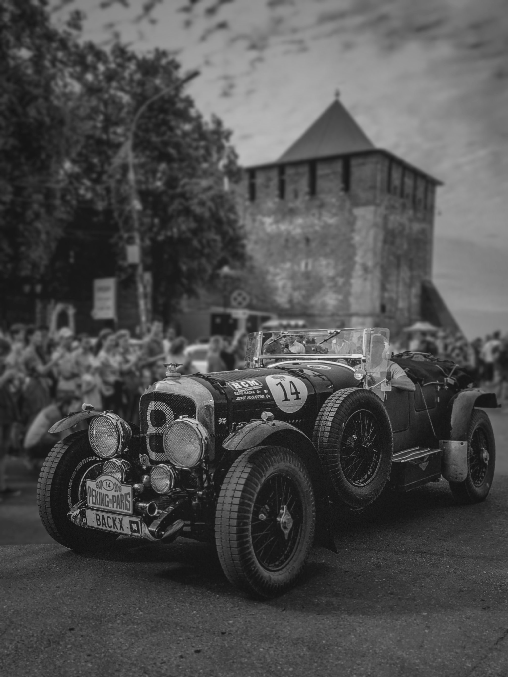 Classic vintage car parked in front of a historic tower, surrounded by an enthusiastic crowd. The monochrome effect enhances the nostalgic atmosphere.