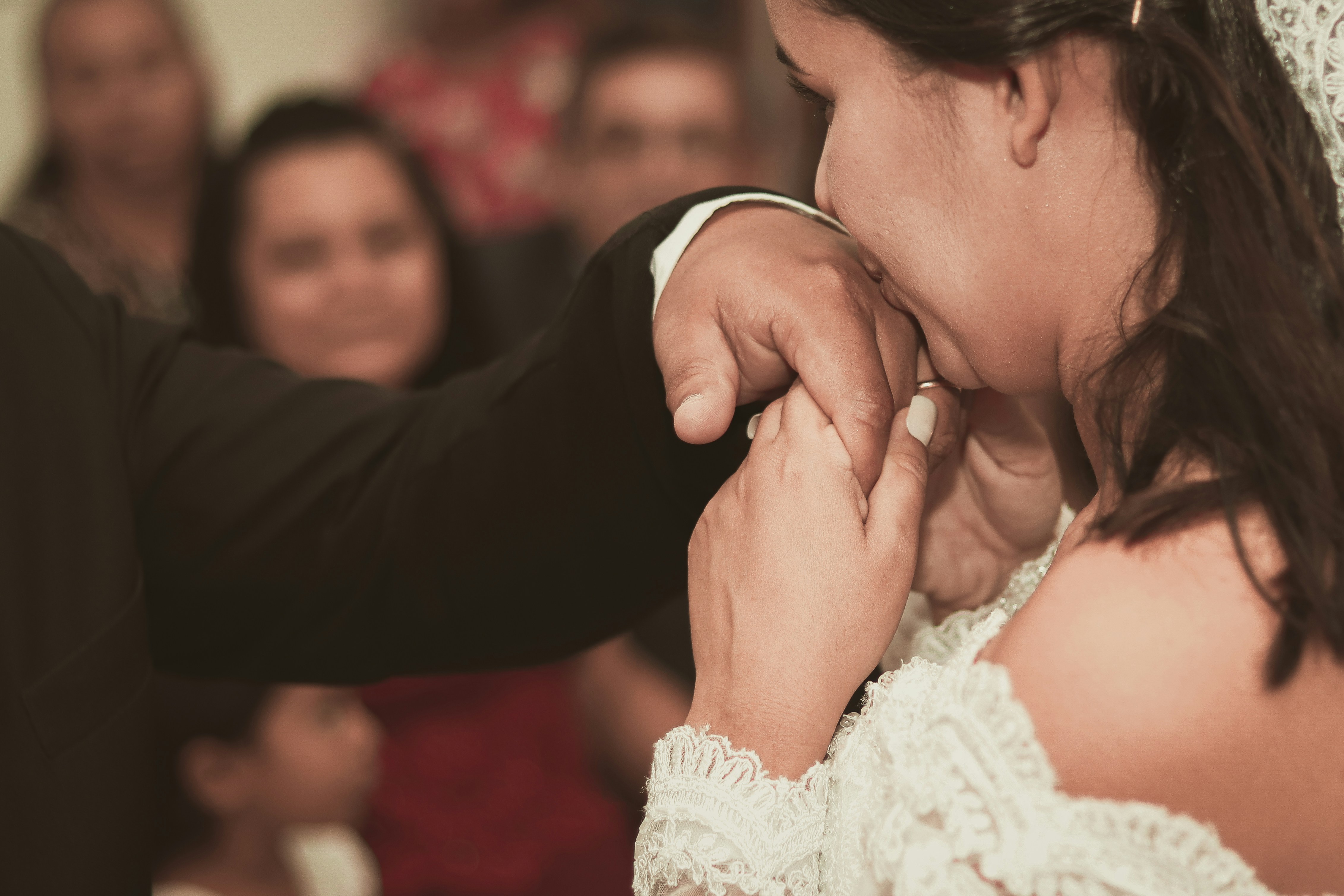 Groom gently kisses the bride's hand during a wedding ceremony, surrounded by softly focused guests.