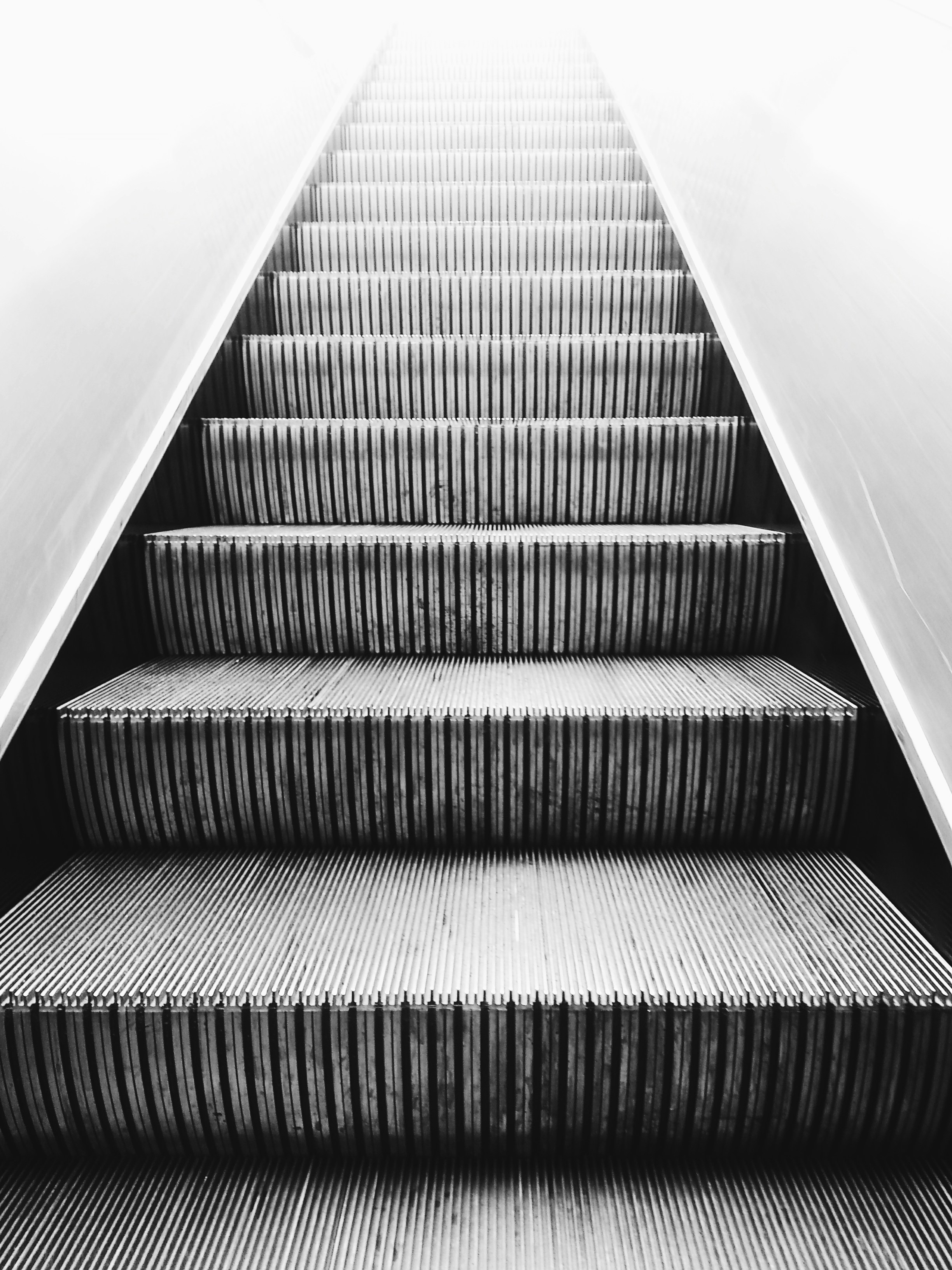 Monochrome photograph of an escalator viewed from the base, emphasizing repeating steps and converging lines.