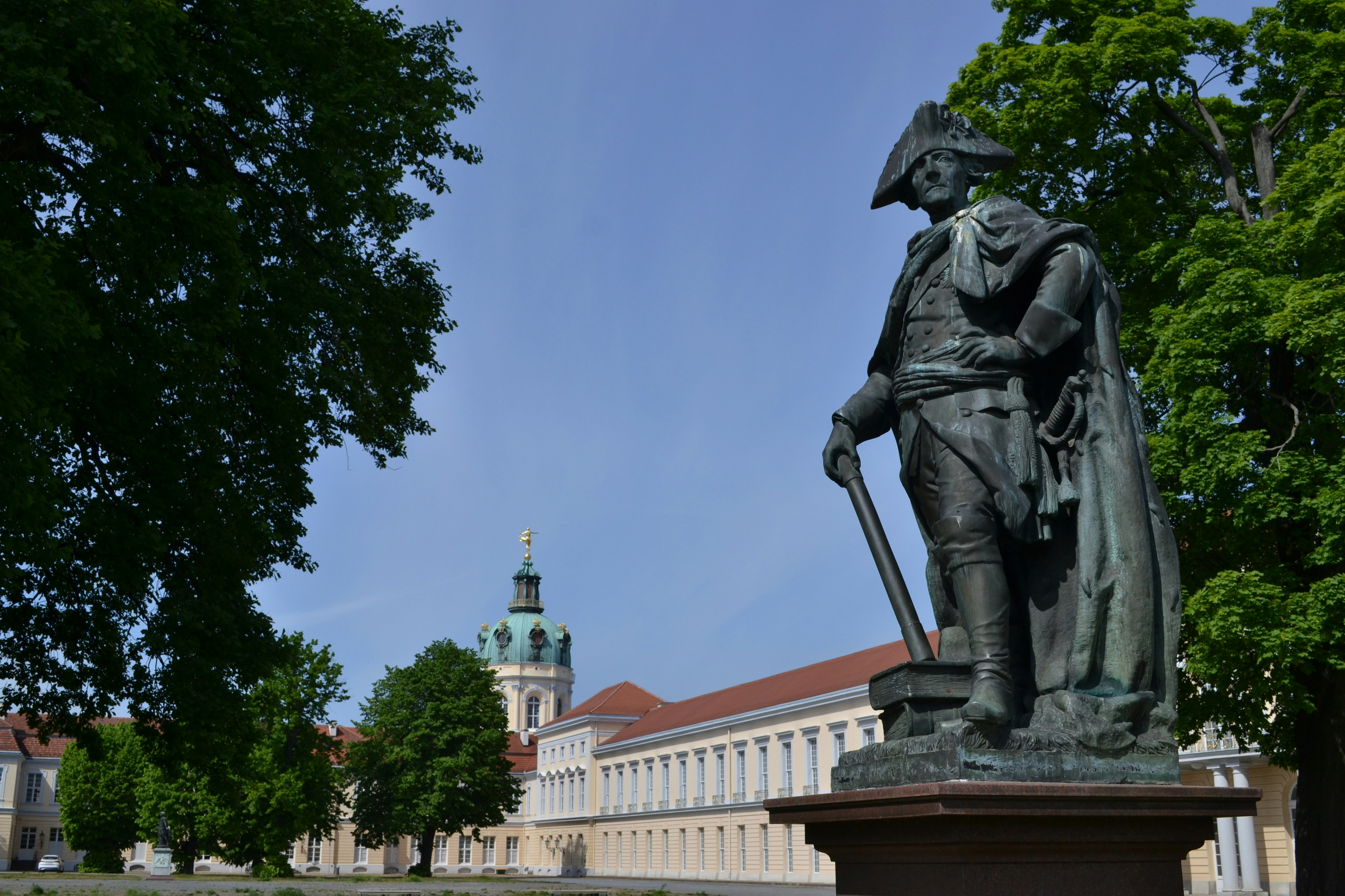 Bronze statue of a historical figure standing majestically, framed by lush greenery and classic architecture in the background.
