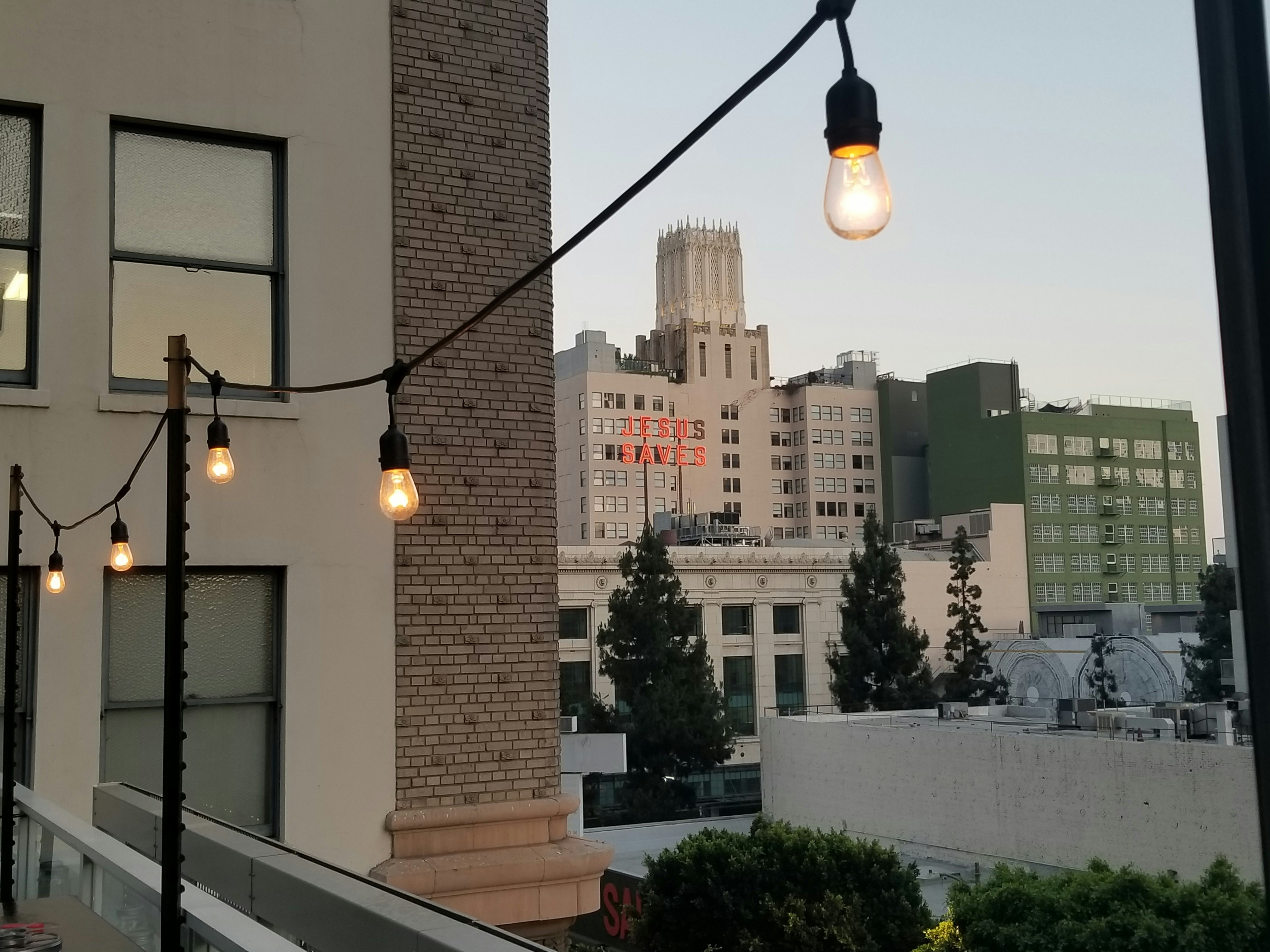 String lights illuminate a balcony overlooking a cityscape featuring historic architecture and modern buildings. The scene captures the essence of urban living at dusk.