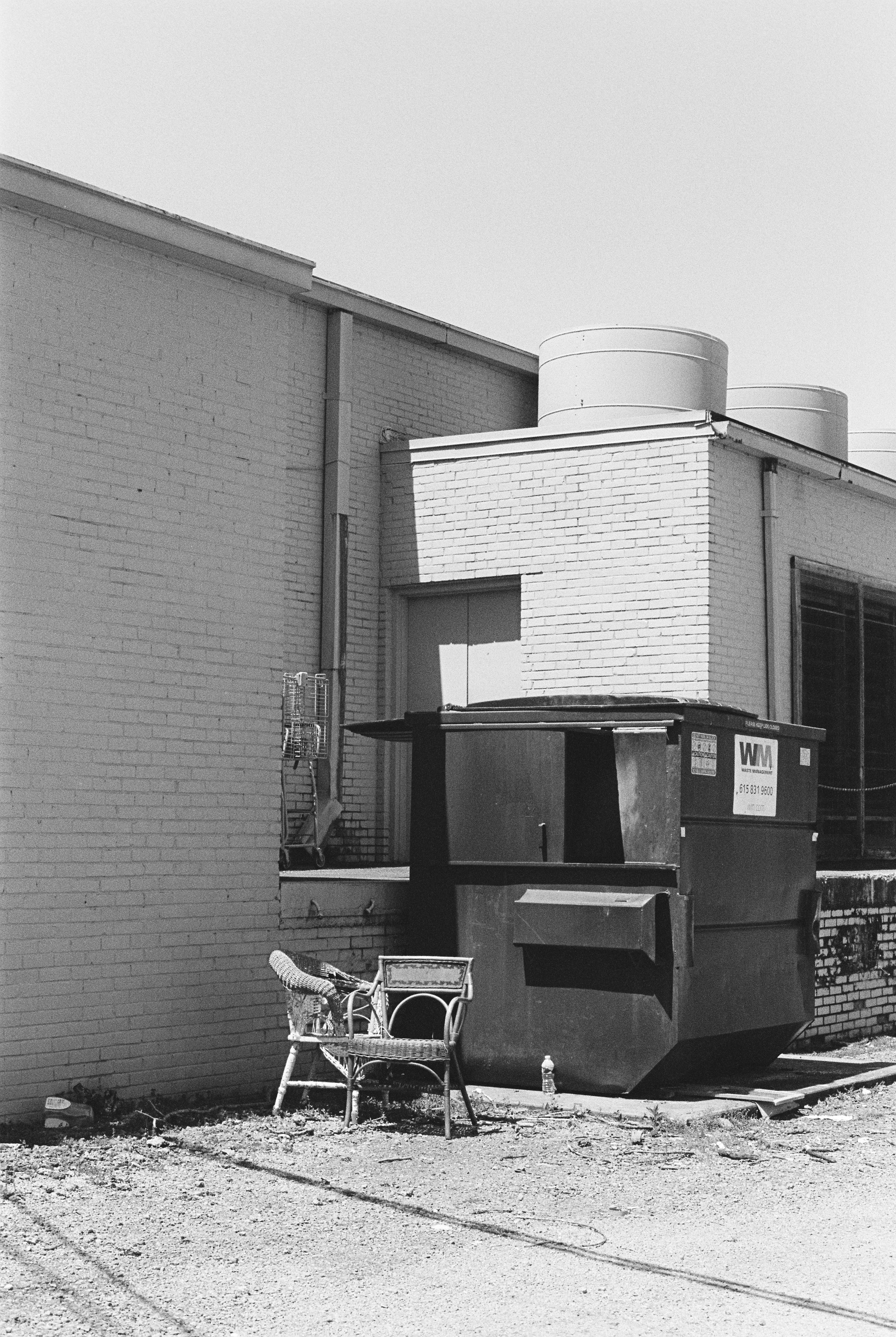 A black and white photograph showcasing a dumpster beside a building, with a discarded chair nearby, highlighting themes of neglect and urban decay.