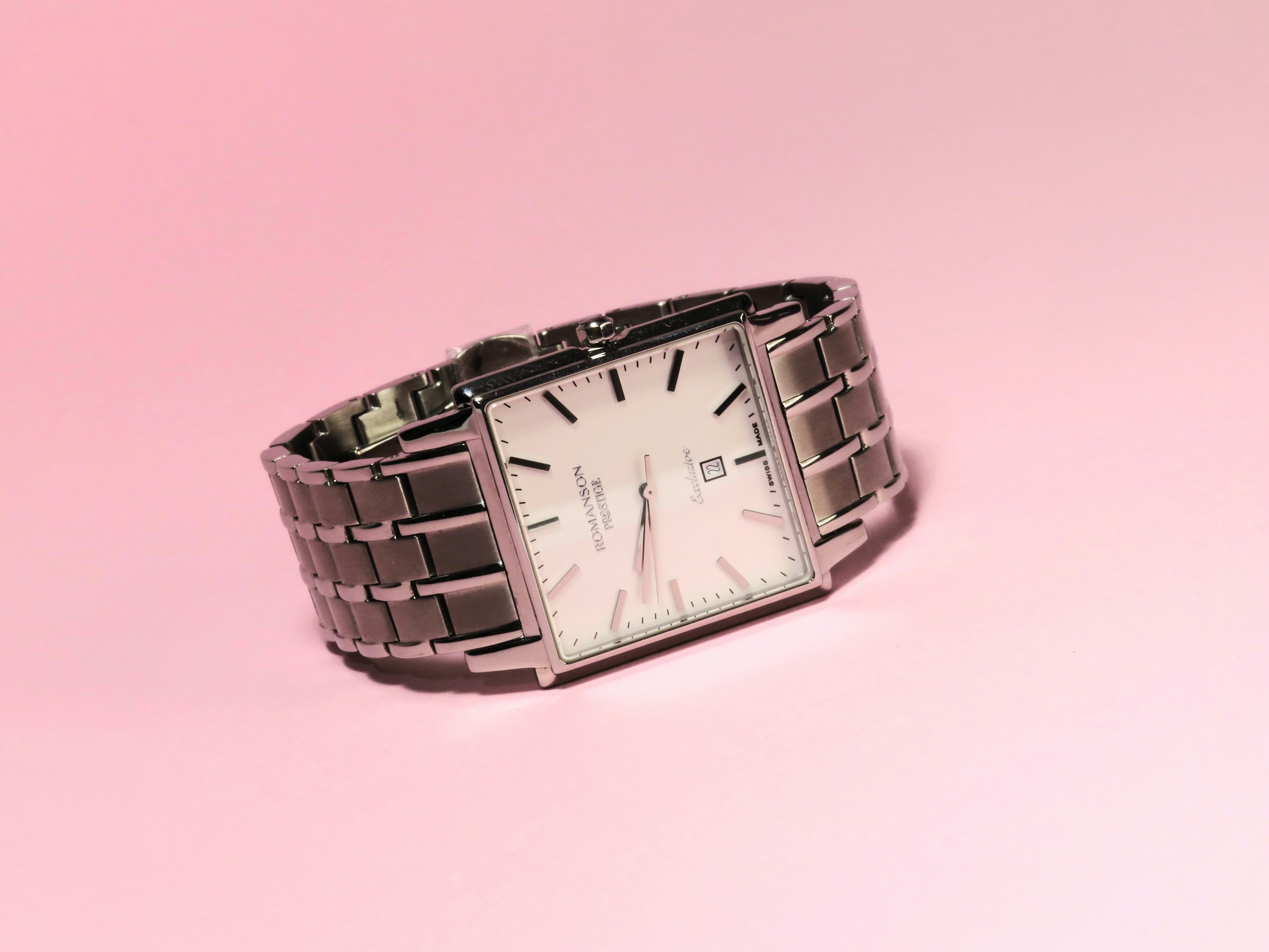 Silver stainless steel wristwatch rests on a pastel pink surface, angled to reveal the rectangular dial. The shot emphasizes minimalist lines and metal finish.