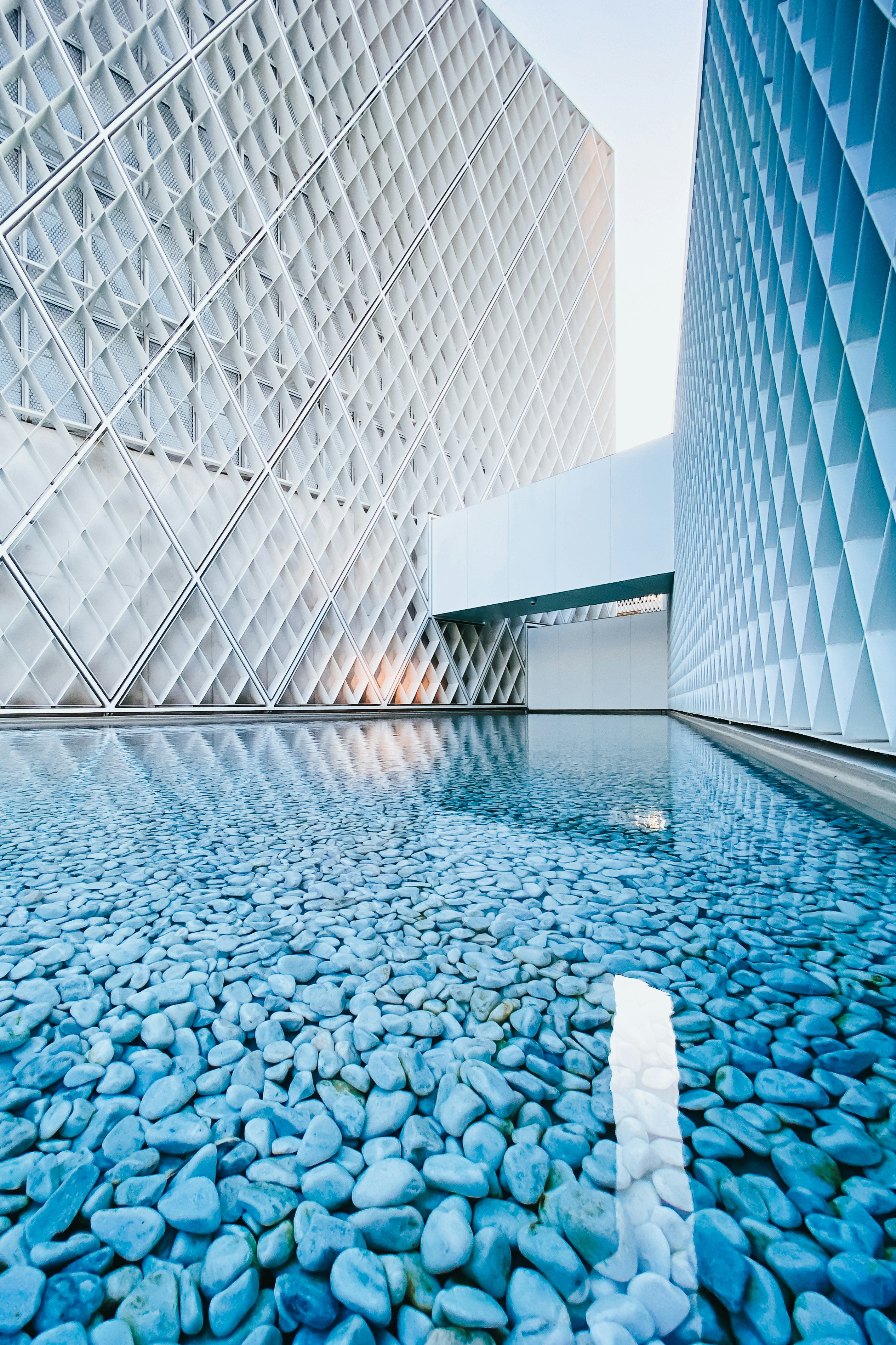 Architectural photograph of diamond-patterned facades flanking a reflective pool filled with blue pebbles.