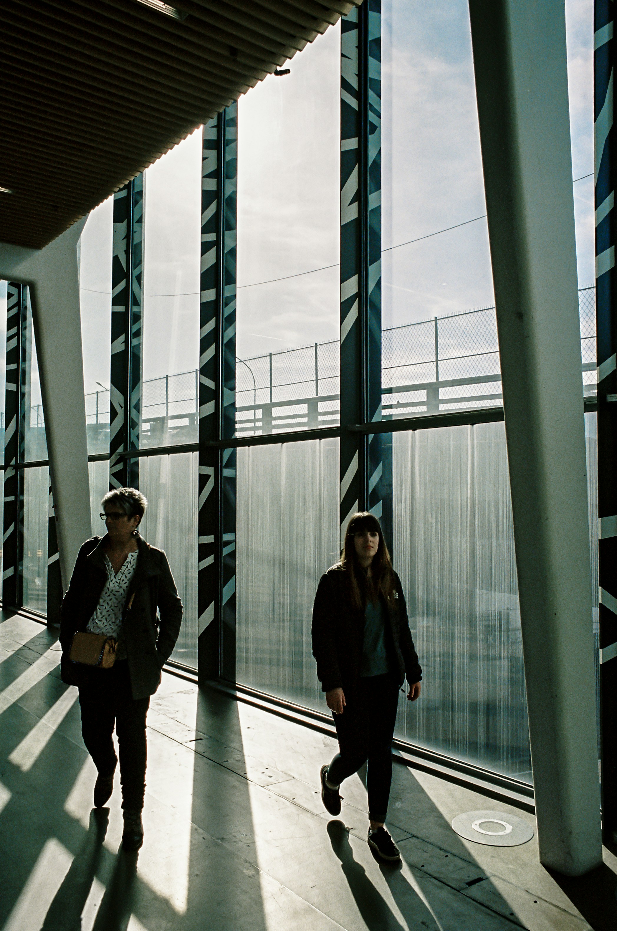 Footpath Between Glass Veins: Two Travelers in a Sunlit Corridor