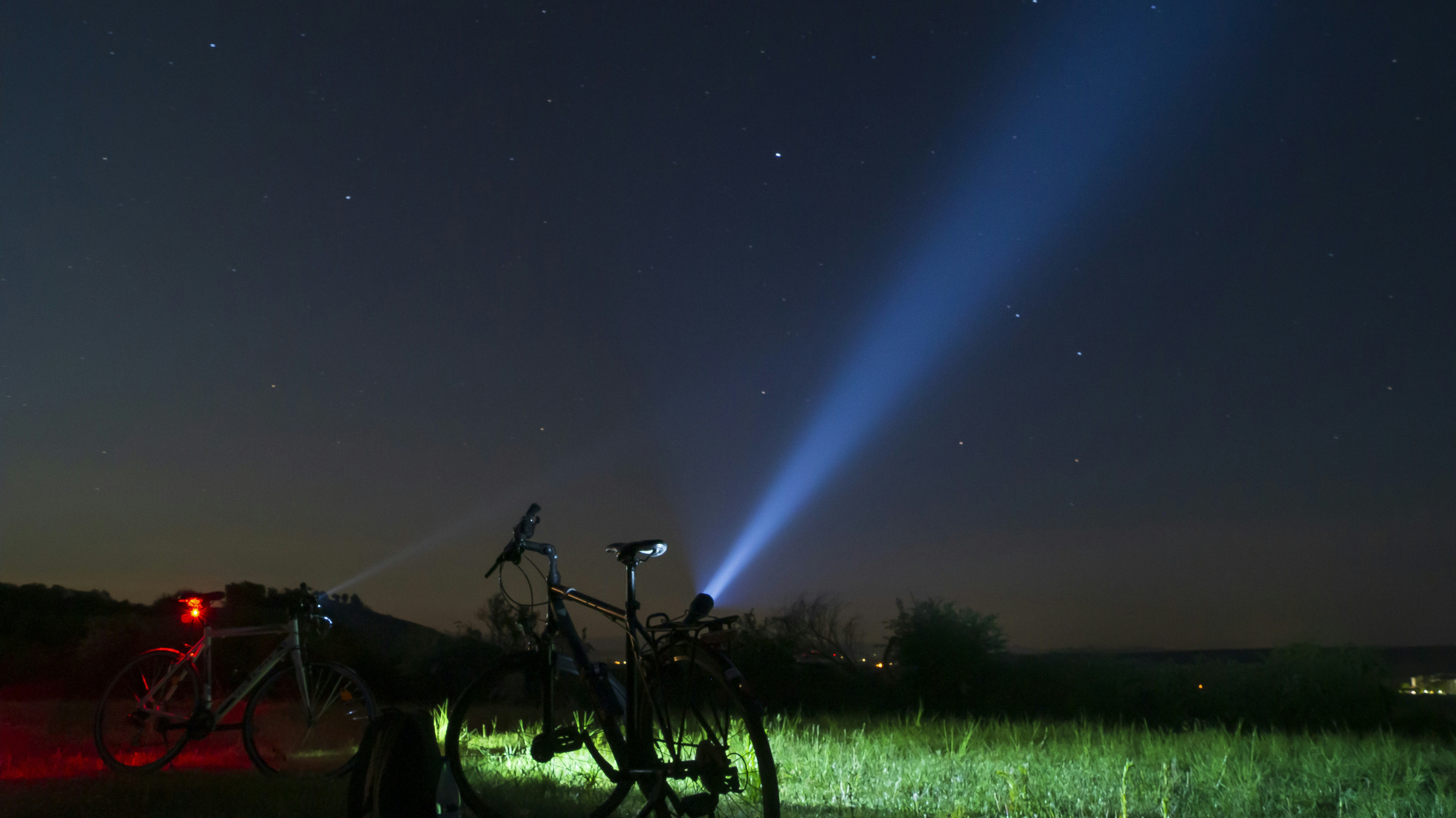 Two bicycles stand on a grassy field under a starry sky, with a powerful flashlight beam cutting through the darkness.