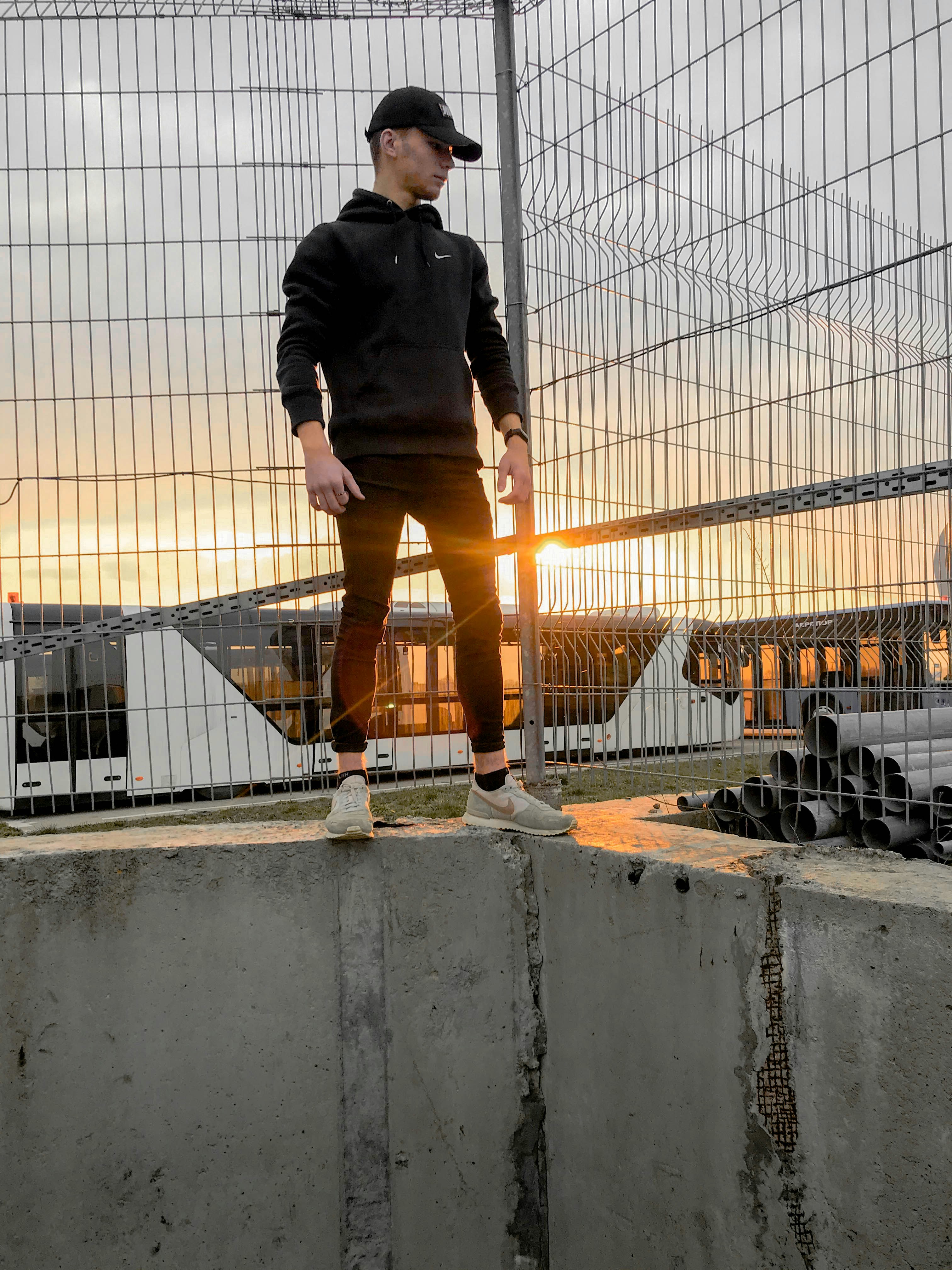 A young man stands on the edge of a concrete structure at sunset, framed by a chain-link fence with industrial elements in the background.