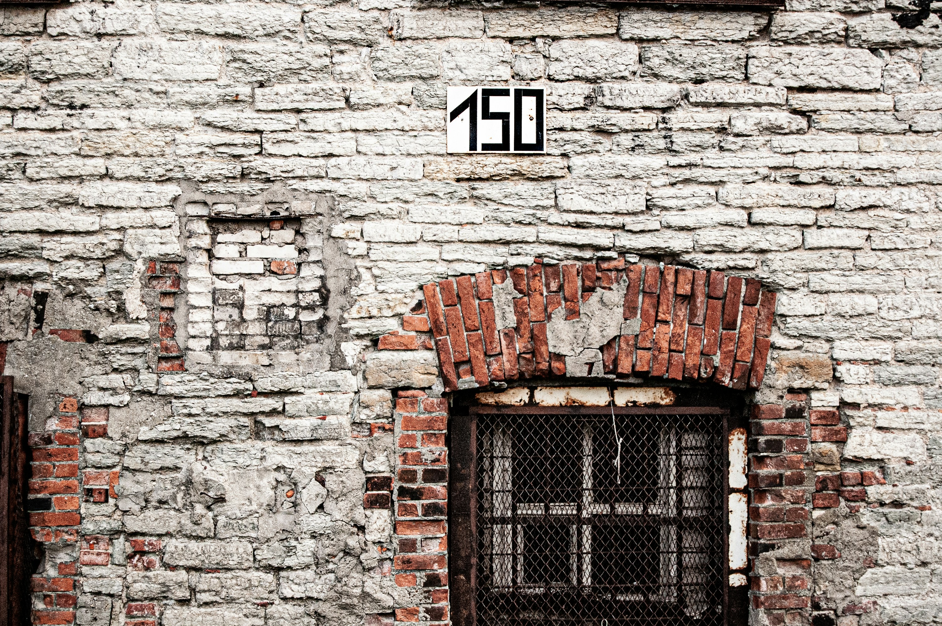 Photograph of a weathered brick wall featuring a brick-arched doorway with a metal grate window. A white '150' sign sits above, creating a stark focal point.