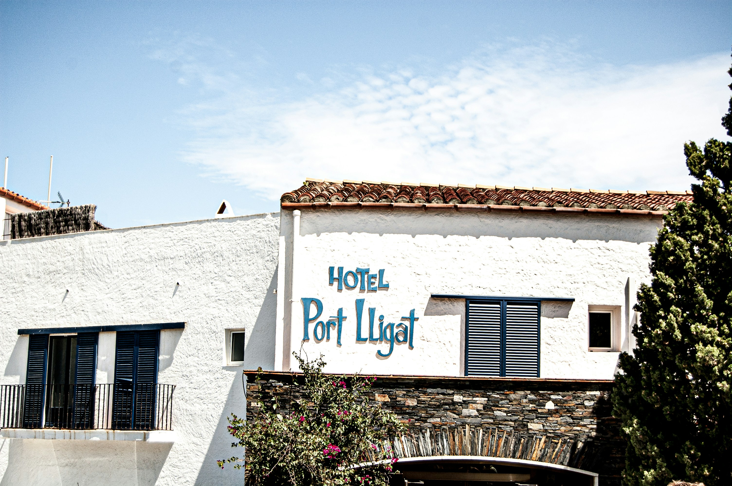 Hotel Port Lligat showcases a whitewashed facade with vibrant blue lettering, framed by lush greenery. The inviting structure reflects a coastal aesthetic.