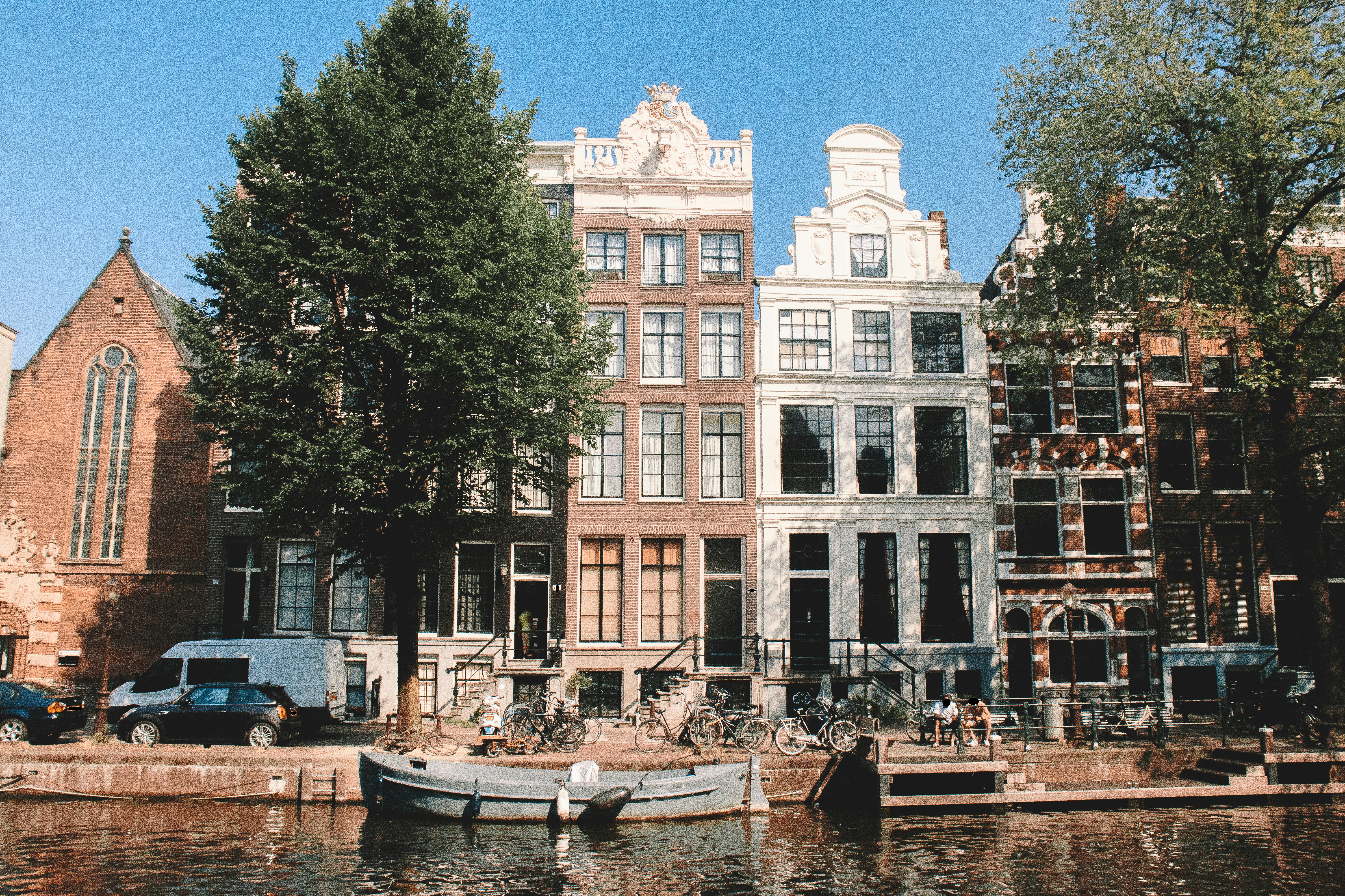 Row of historic buildings lining a serene canal, showcasing a blend of architectural styles with bicycles and a boat in the foreground.