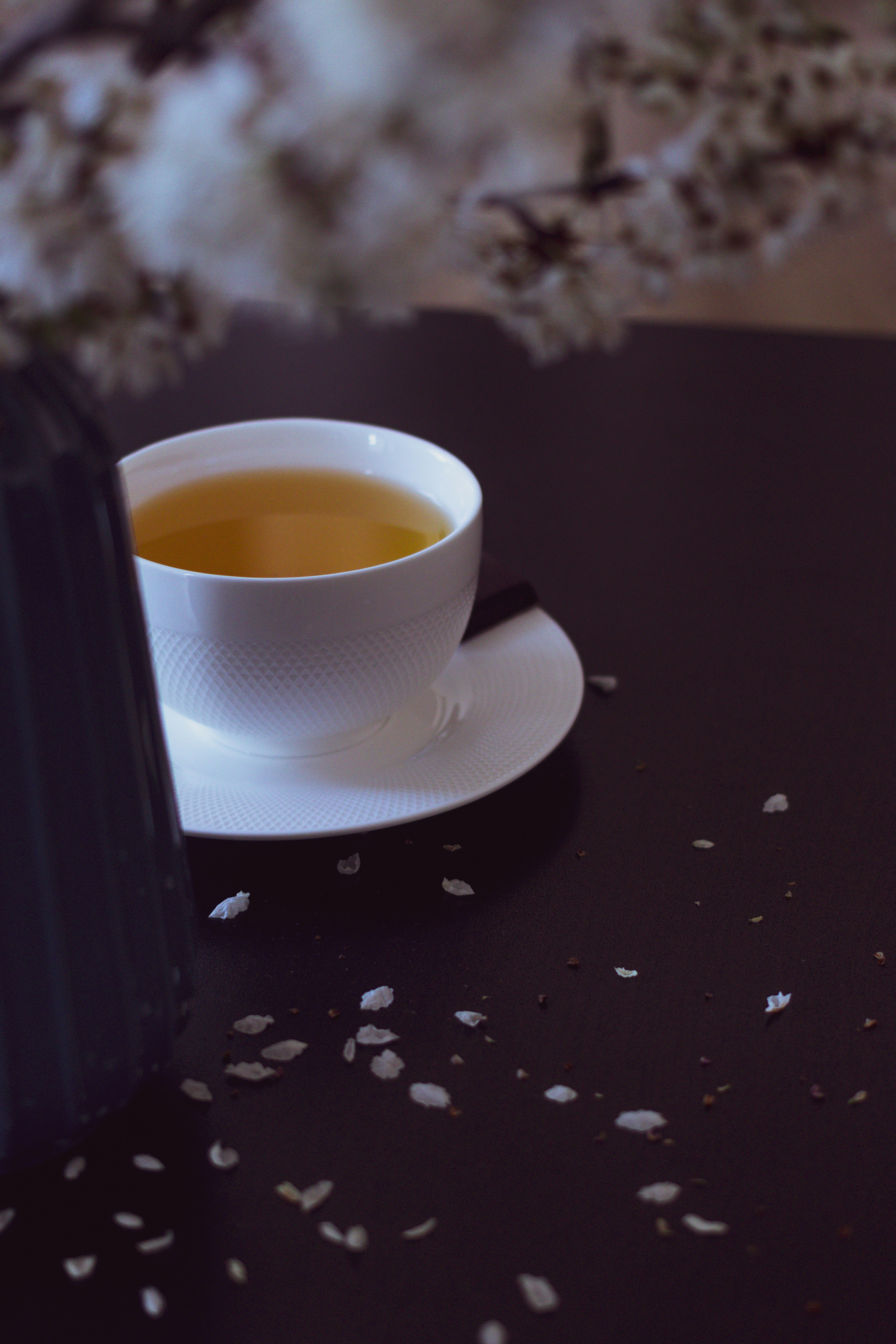 Delicate white tea cup filled with amber tea, accompanied by scattered petals and a floral backdrop. A tranquil setting for relaxation.