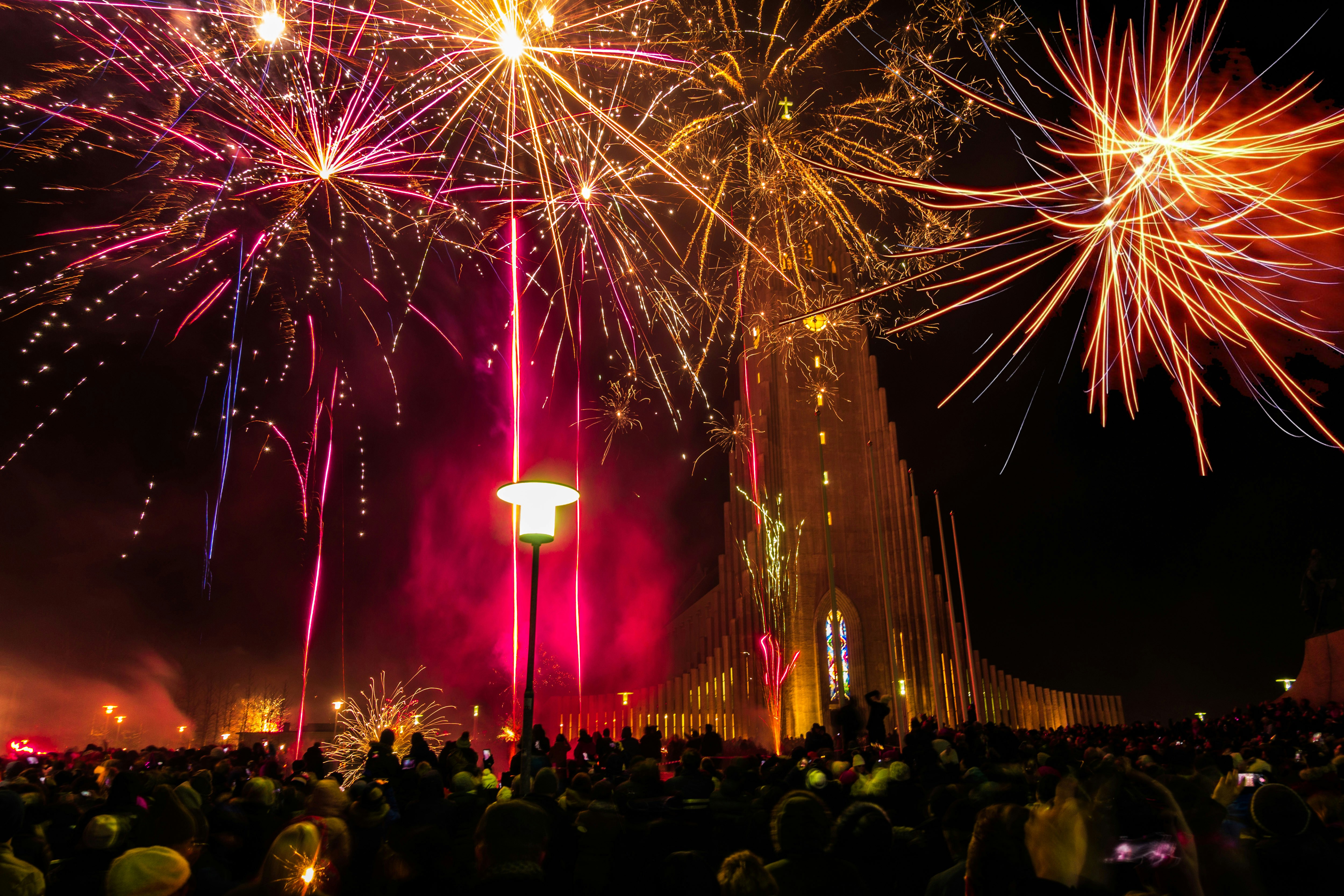 Fireworks burst over a crowd gathered near a brightly lit architectural structure at night.