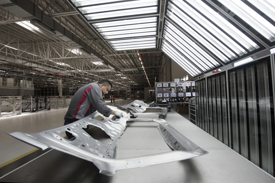 Photograph of a technician inspecting aluminum car body panels on a production line beneath skylights. The scene highlights a high-tech manufacturing environment with racks and glass-front cabinets in the background.