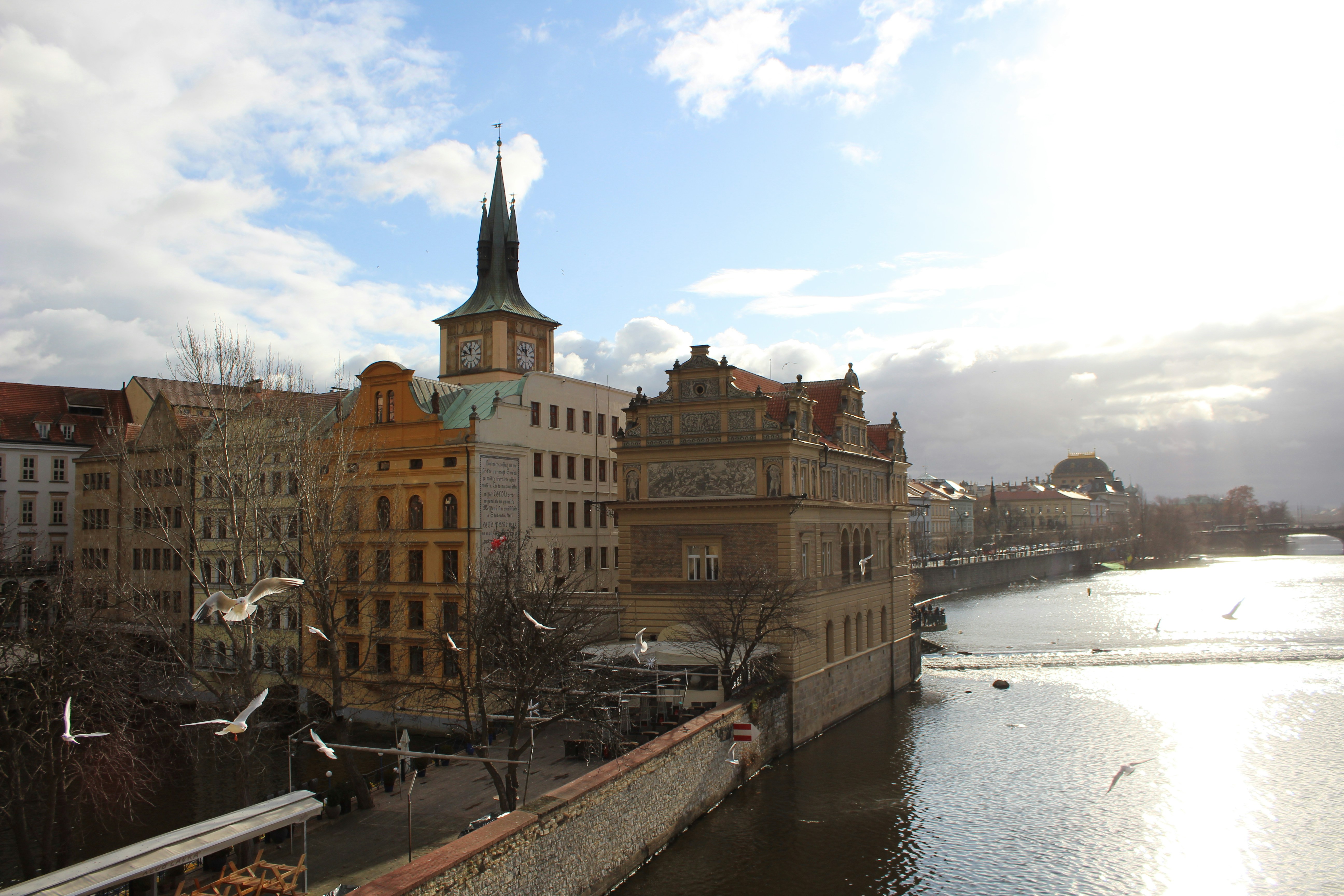 Historic riverside buildings with a prominent spire under a bright sky.