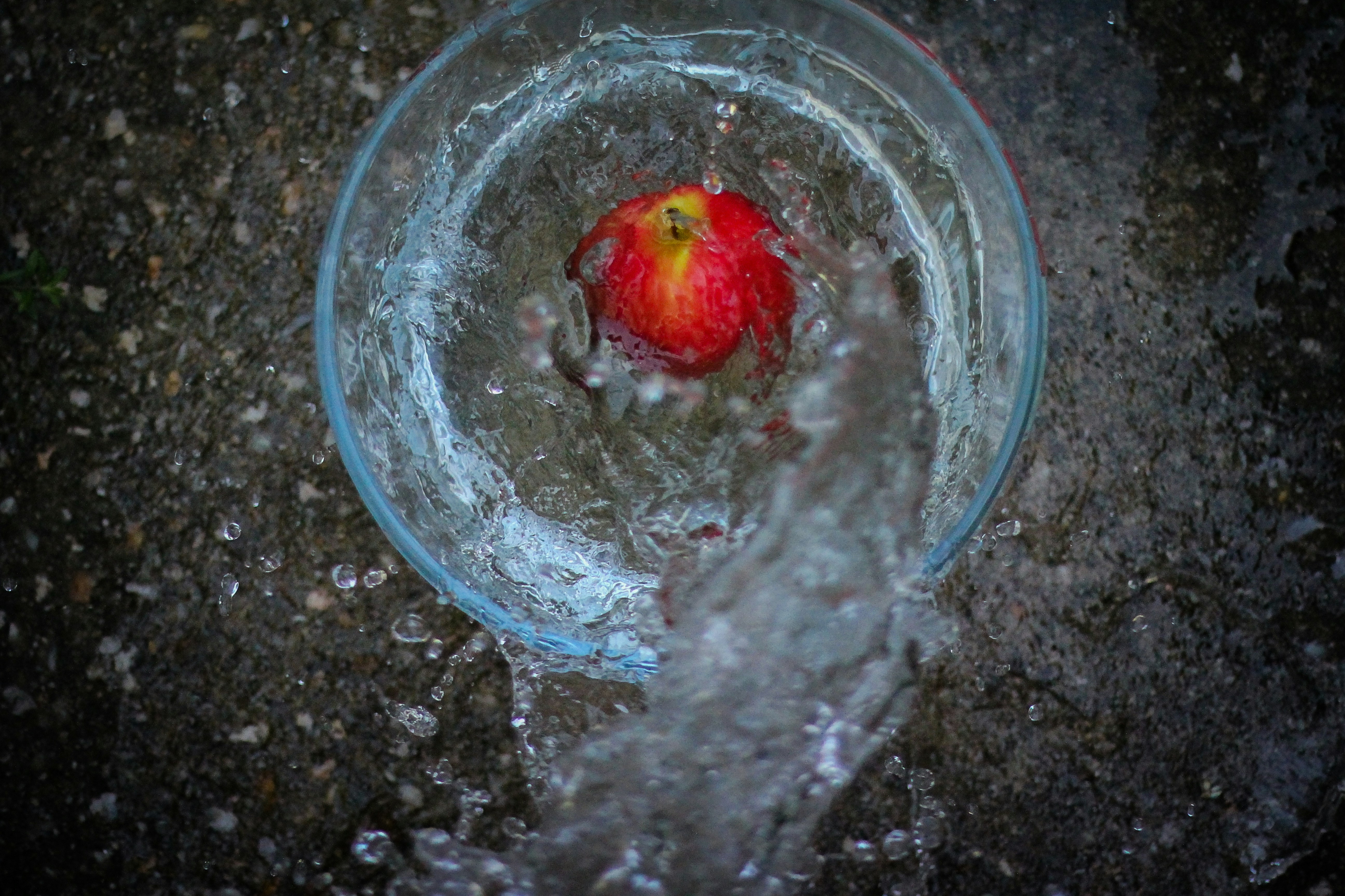 A vibrant red apple splashes into a clear bowl of water, creating dynamic ripples and droplets. The scene captures the moment of impact and the ensuing water spray.