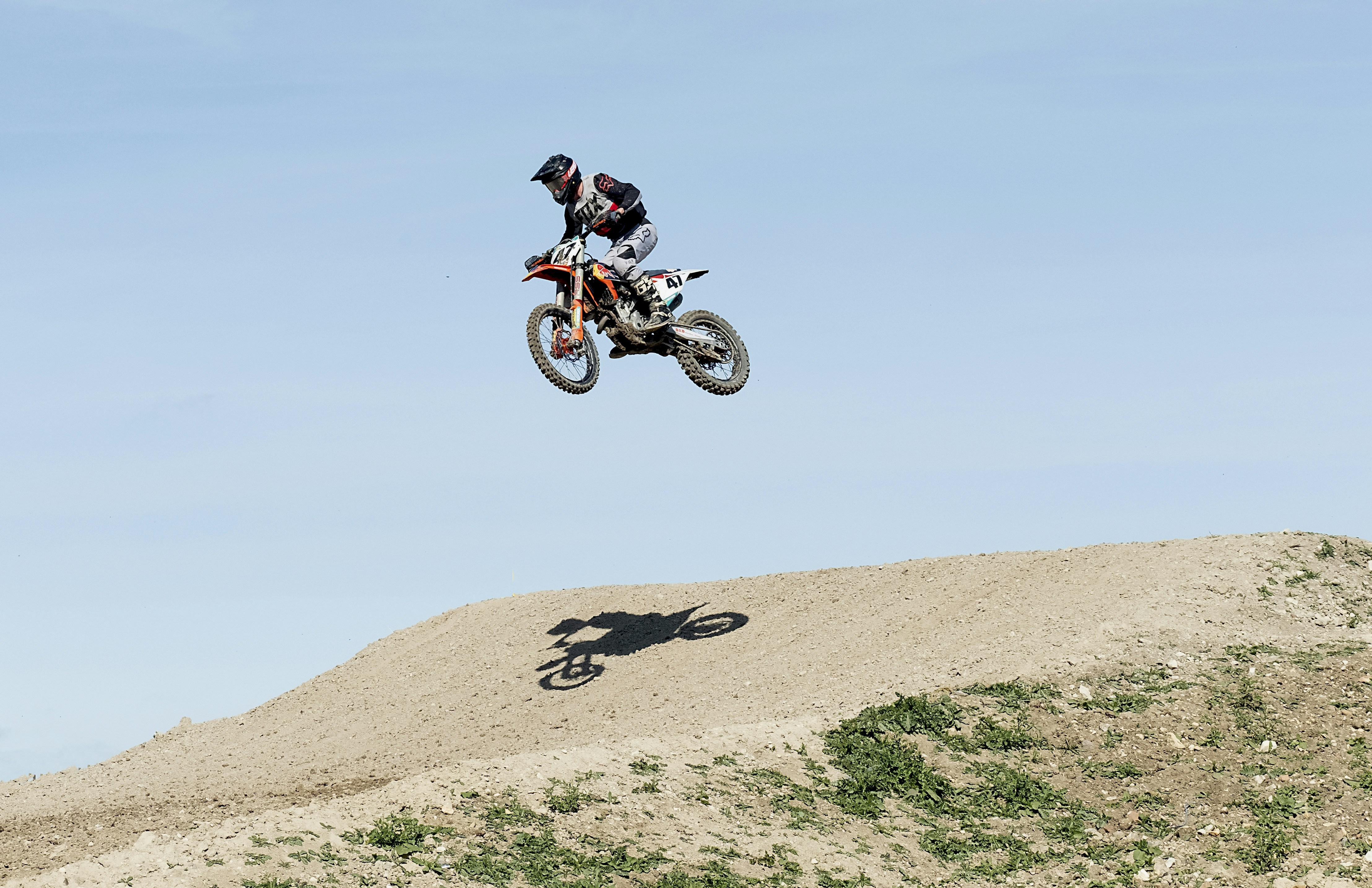 Motocross rider airborne over a dirt track against a clear blue sky.
