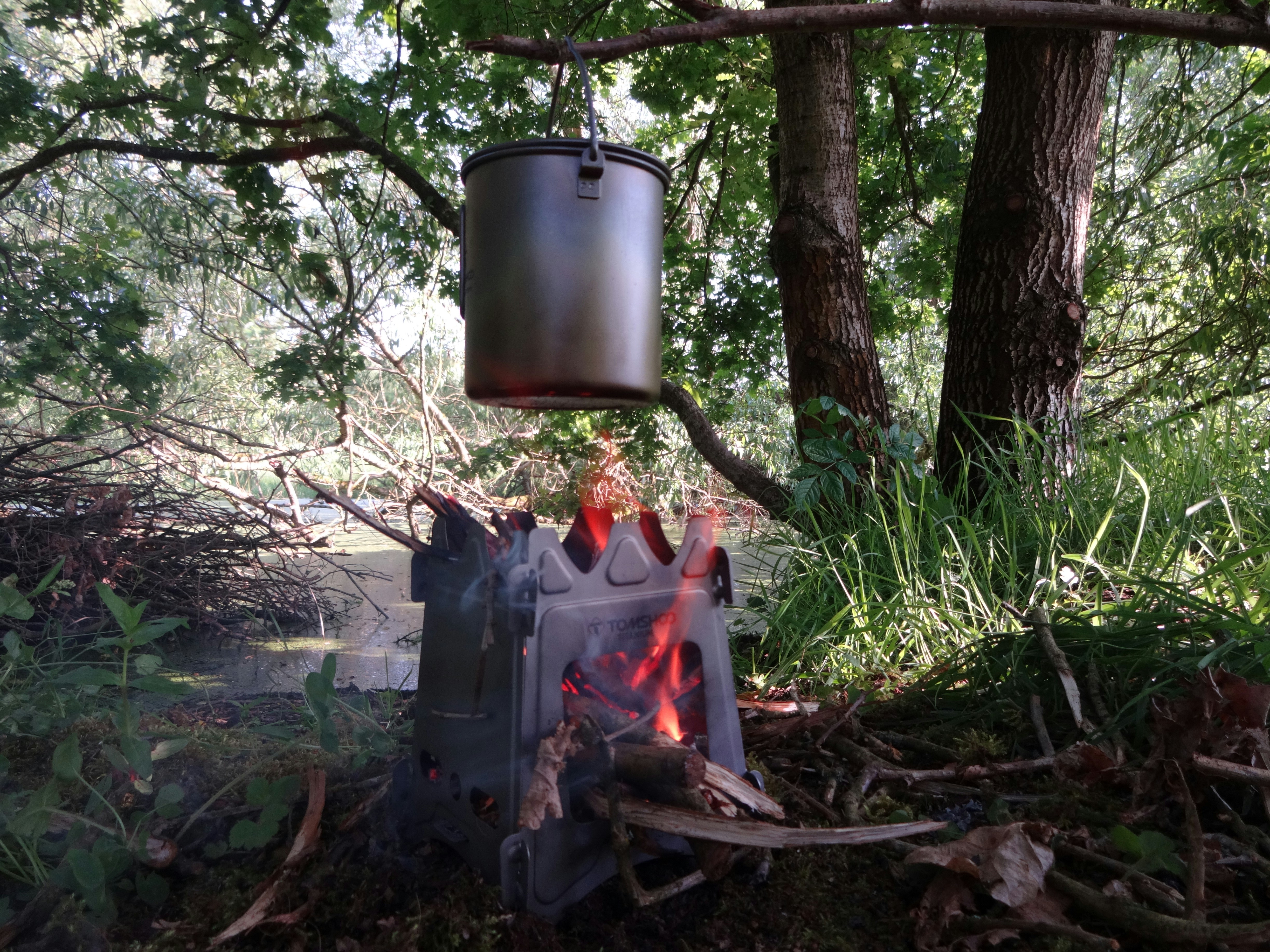 Metal kettle hangs over a compact wood stove in a shaded forest clearing, with embers glowing beneath.