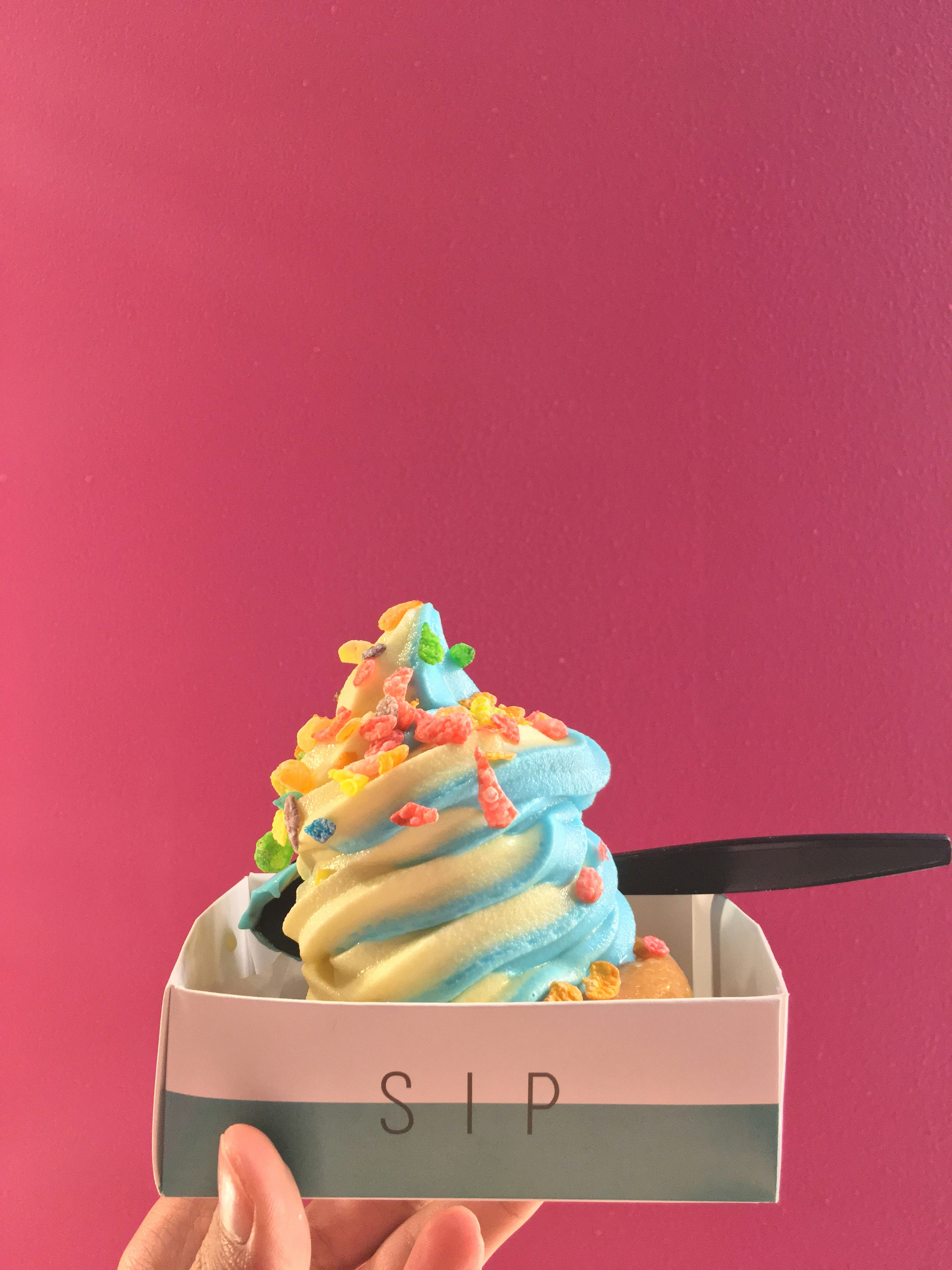 Colorful soft serve ice cream topped with vibrant sprinkles, held against a bright pink backdrop.