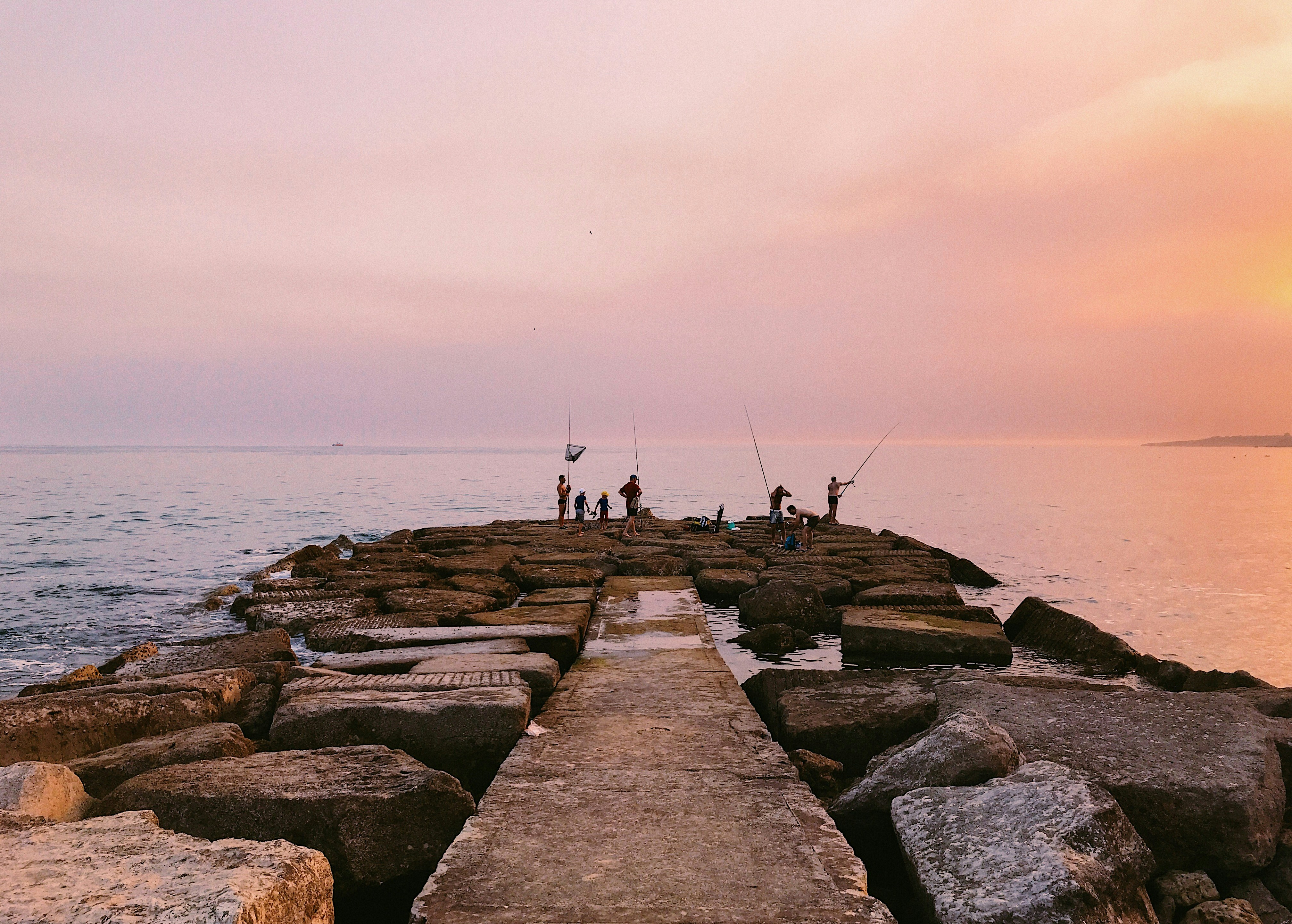 Silhouetted figures fishing on a rocky jetty under a vibrant sunset sky.
